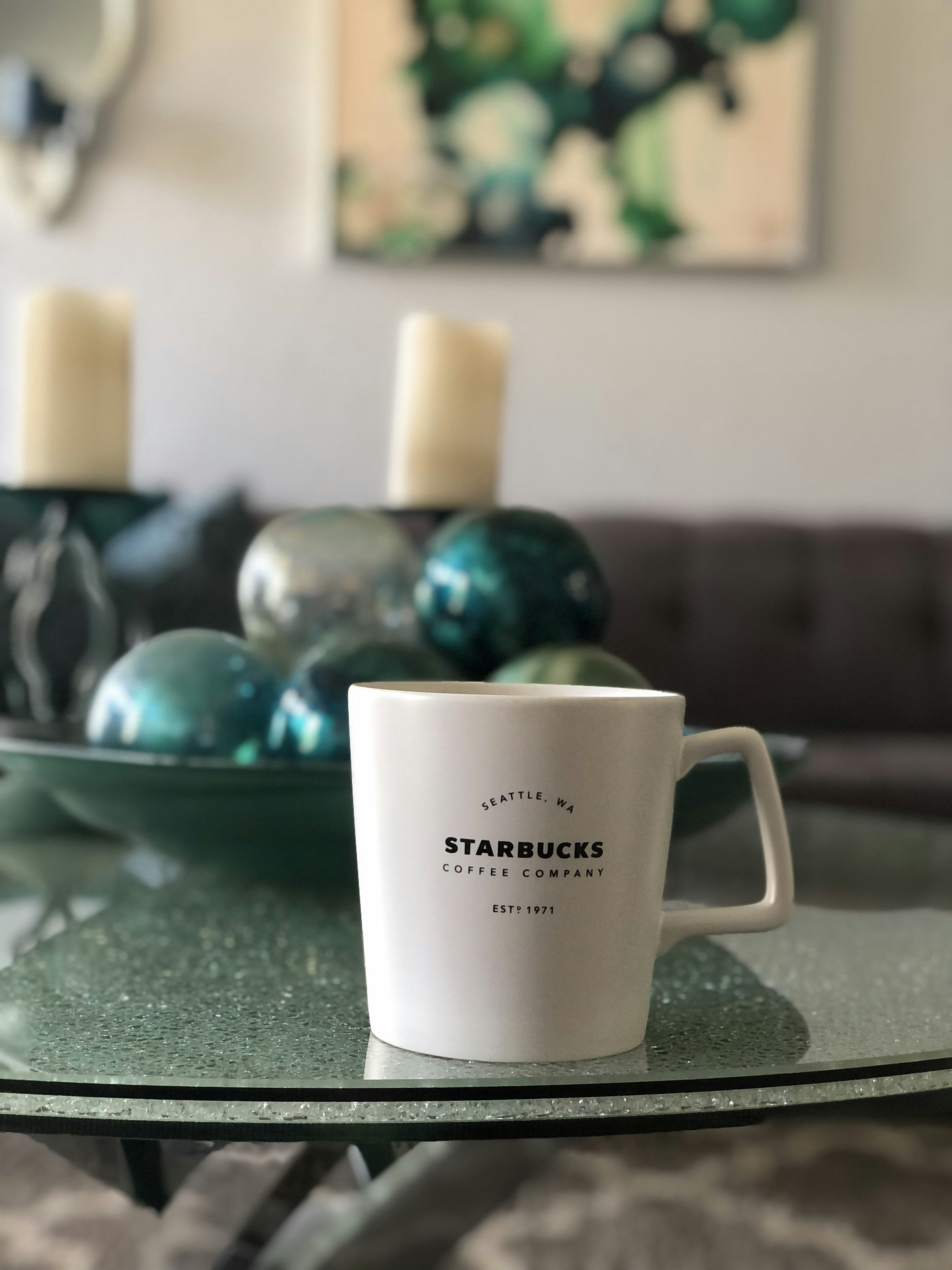 White Starbucks coffee mug resting on a reflective table, surrounded by decorative teal spheres and candles. The background features a vibrant abstract artwork.