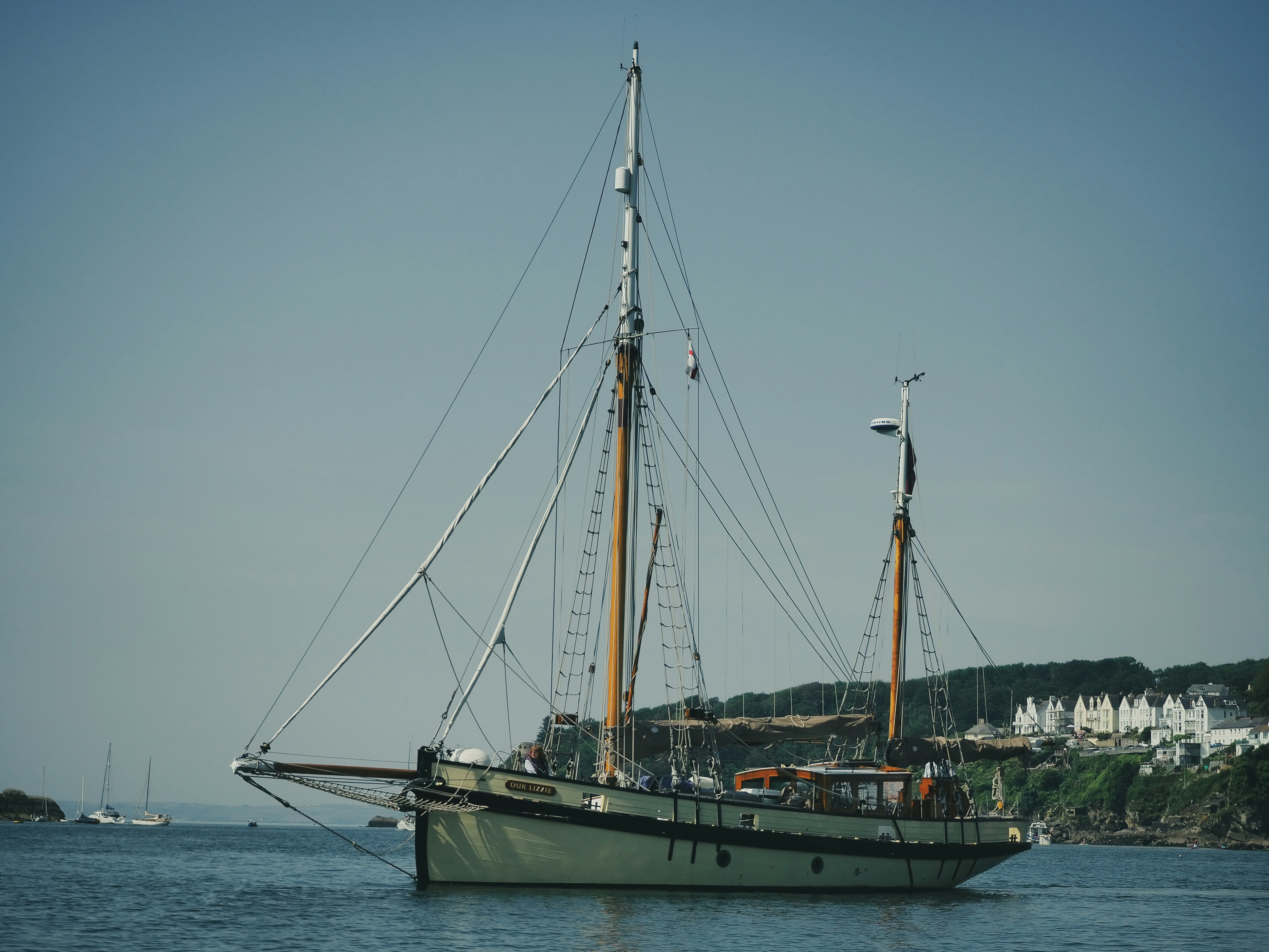 Classic sailing ship anchored in calm waters, with a backdrop of lush green hills and quaint coastal homes.