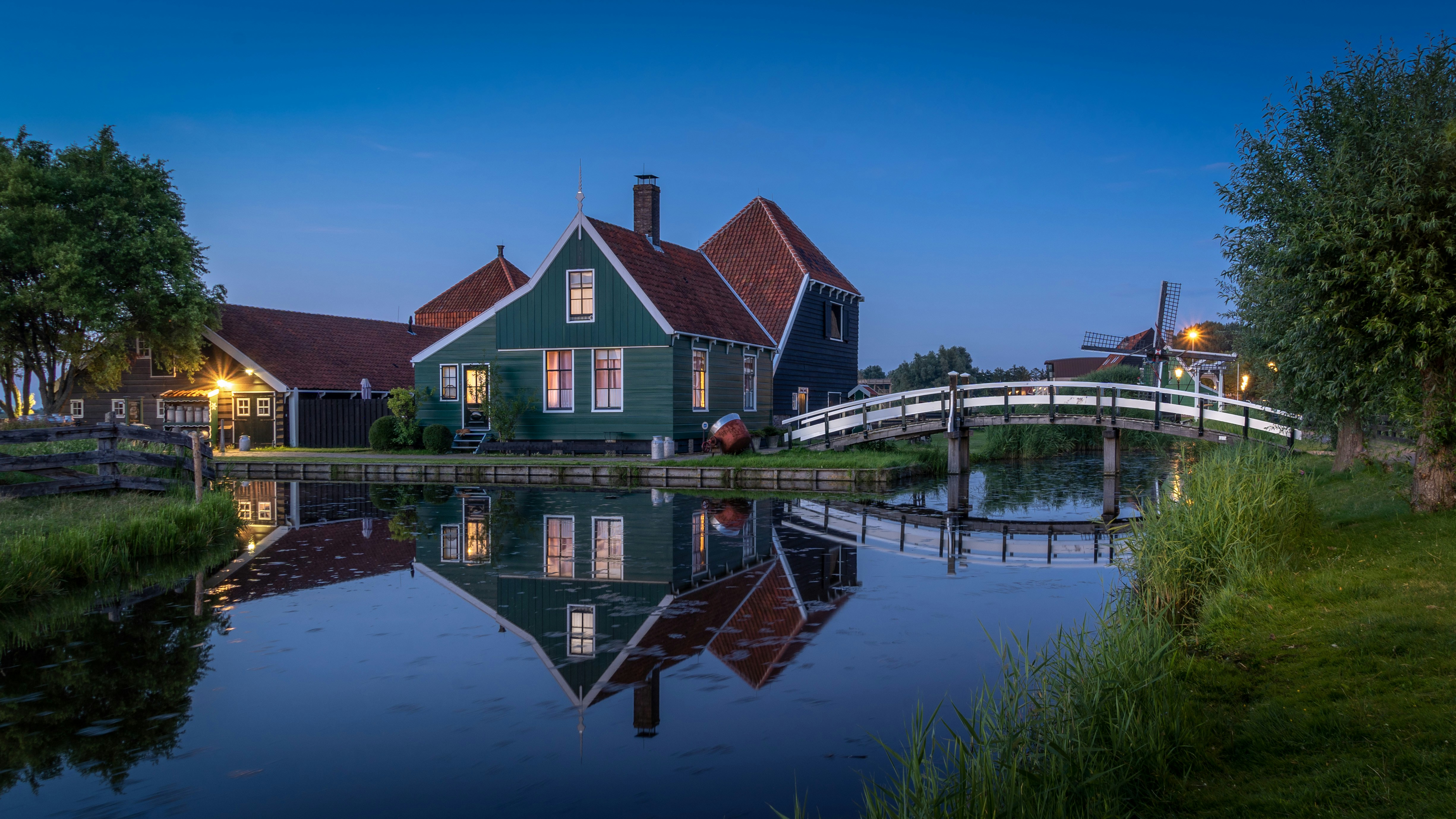 Charming green house with a red roof beside a serene canal, reflecting the twilight sky. A quaint wooden bridge connects the banks, enhancing the peaceful ambiance.