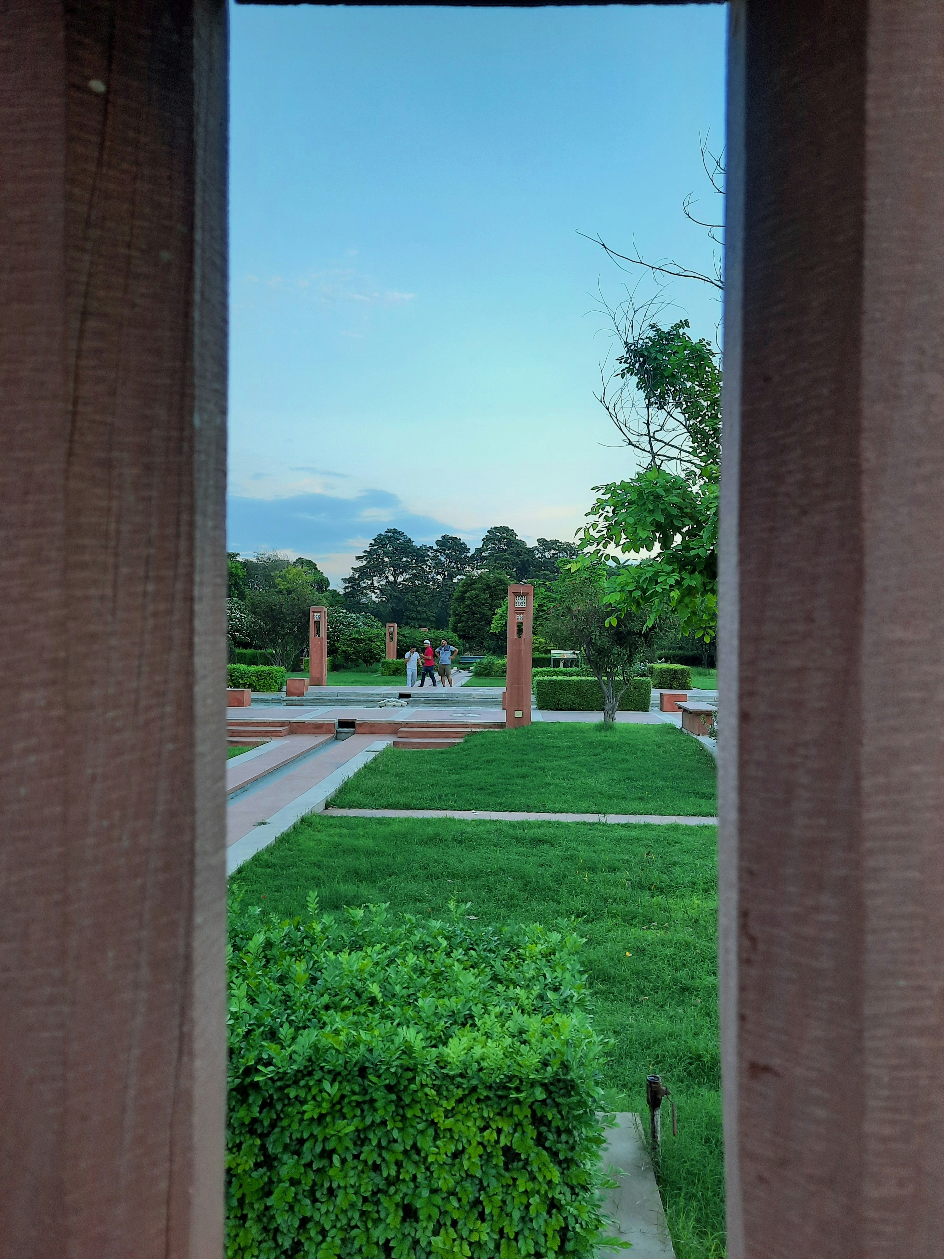 View of a serene park scene framed by wooden structures, showcasing lush greenery and a couple walking along a path.