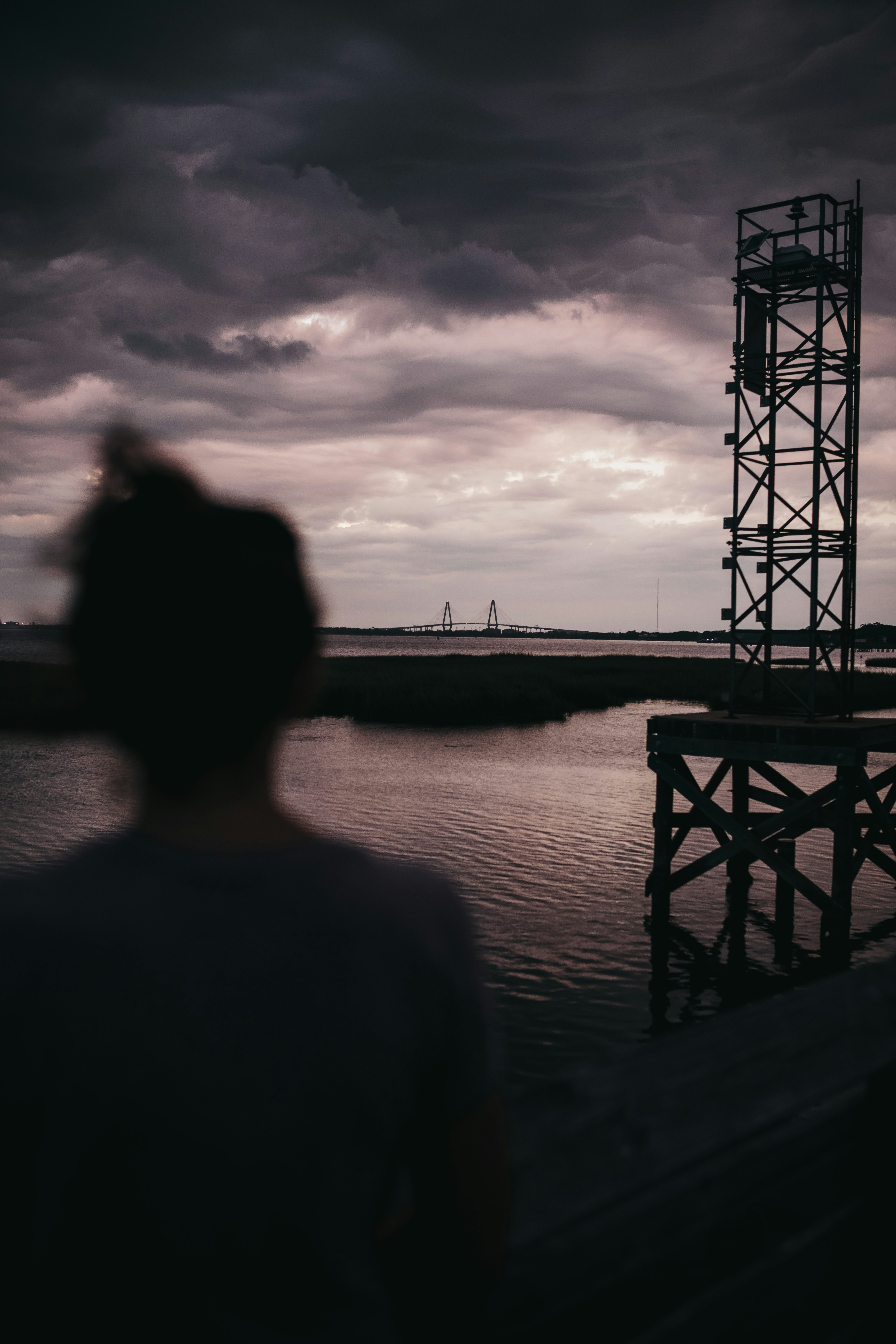 Silhouette of a person overlooking a moody sky and calm waters with a distant industrial structure.