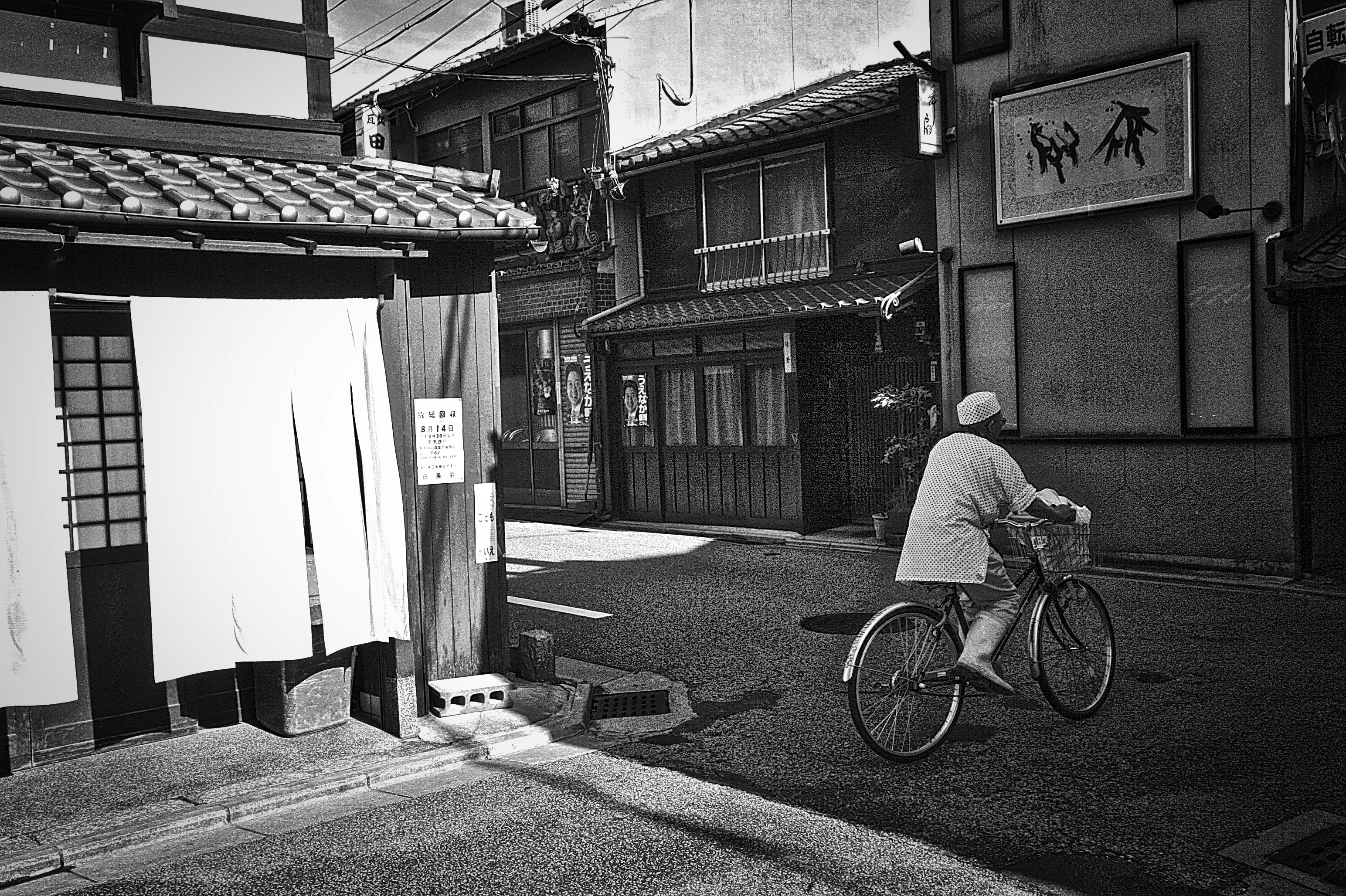 A cyclist in a white outfit rides past traditional Japanese architecture, capturing the essence of daily life in a historic neighborhood.
