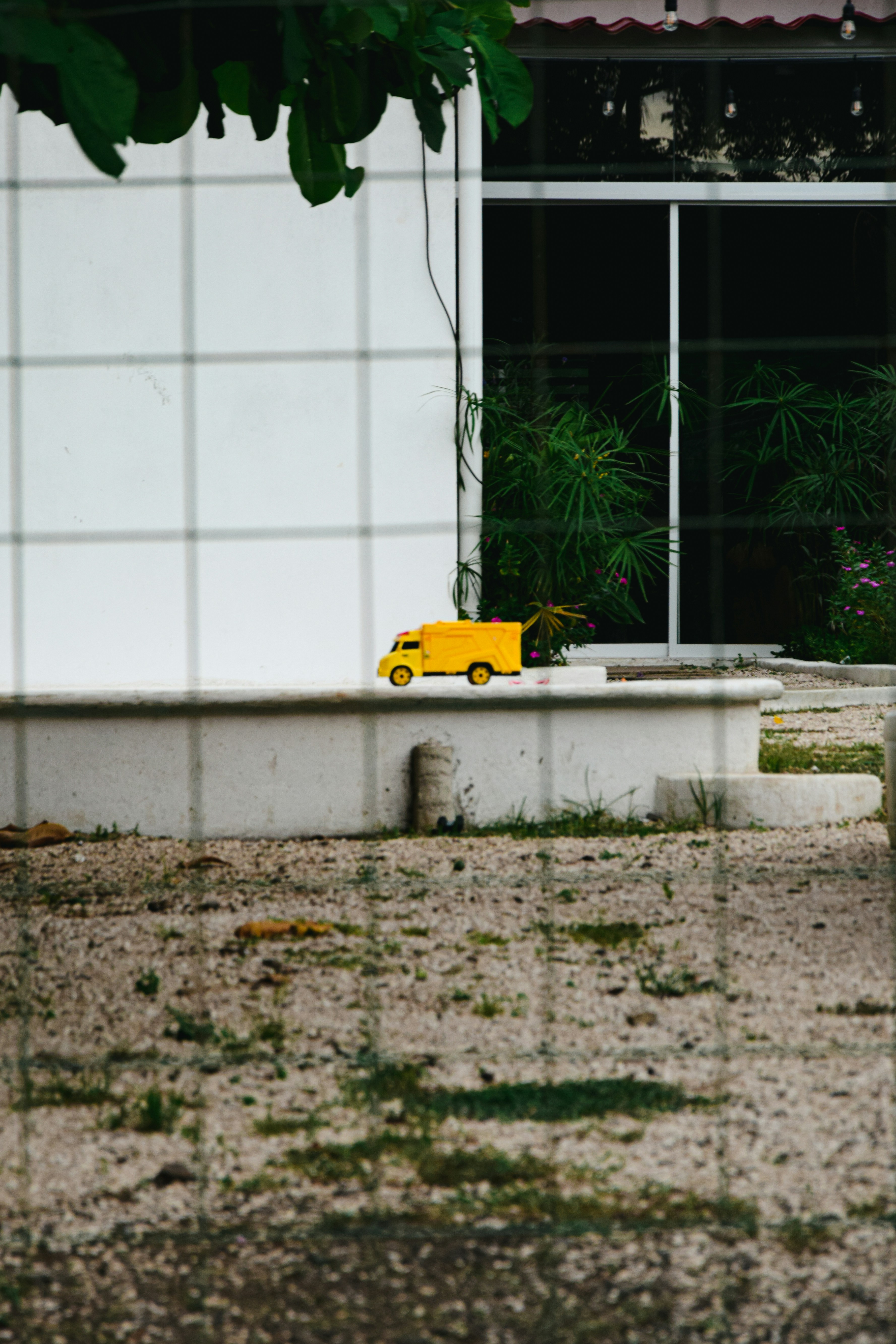 Bright yellow toy truck positioned on a concrete ledge against a backdrop of lush greenery and modern architecture.