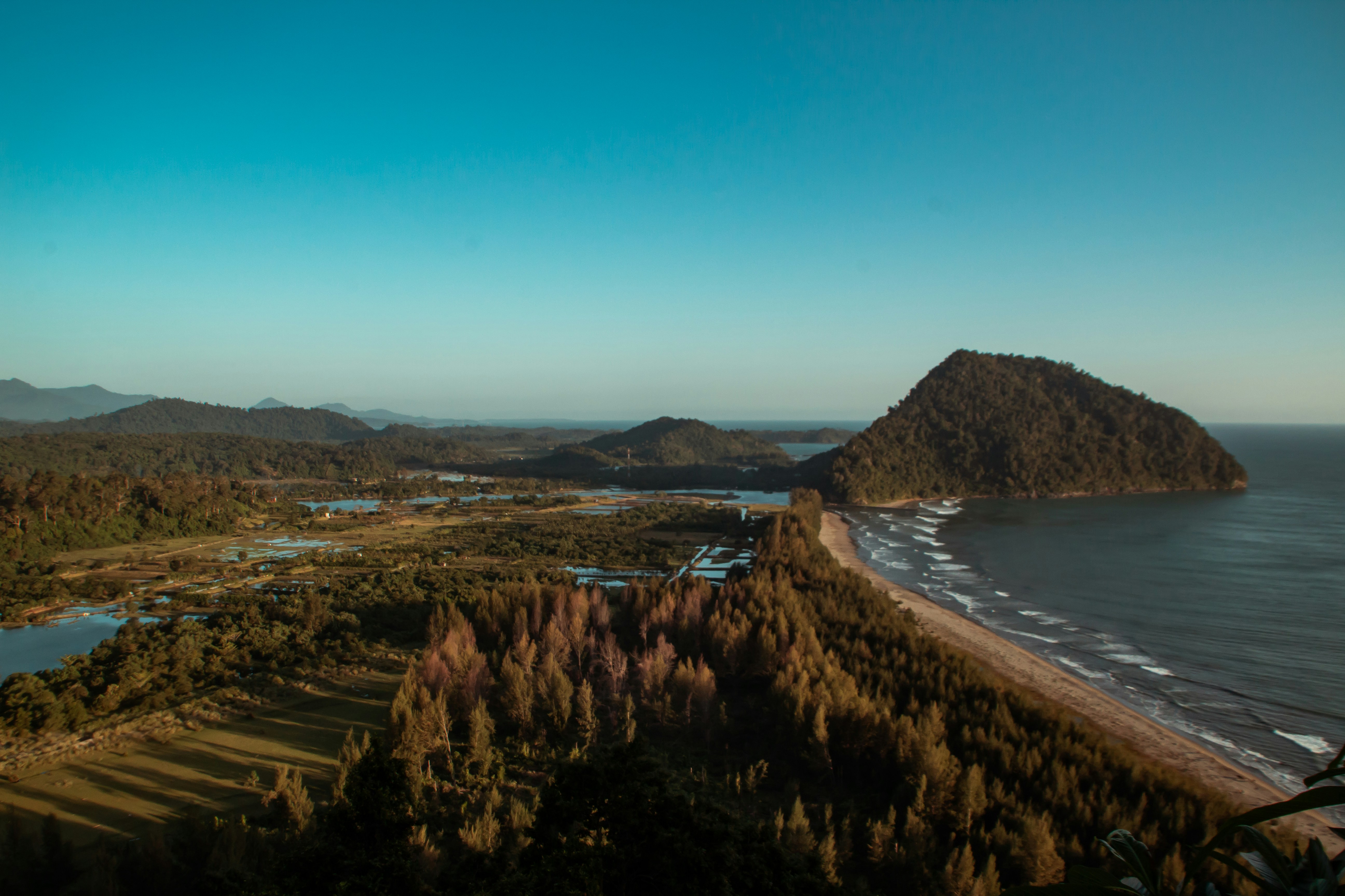 A panoramic view of a tranquil coastal landscape featuring rolling hills, a secluded beach, and a distant mountain silhouette under a clear blue sky.