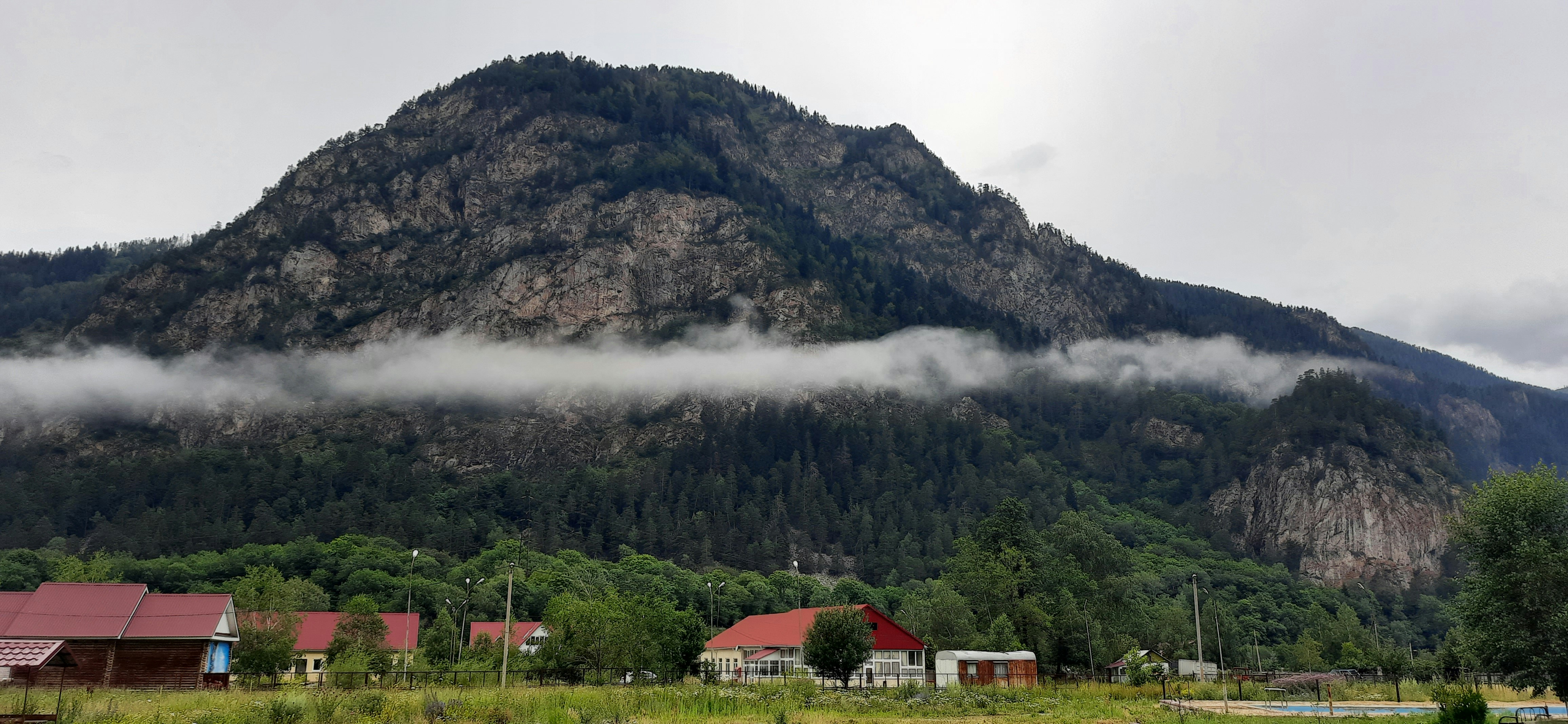Mountain range rises behind a cluster of red-roofed houses in a green valley. A low cloud band sweeps across the mid-mountain.