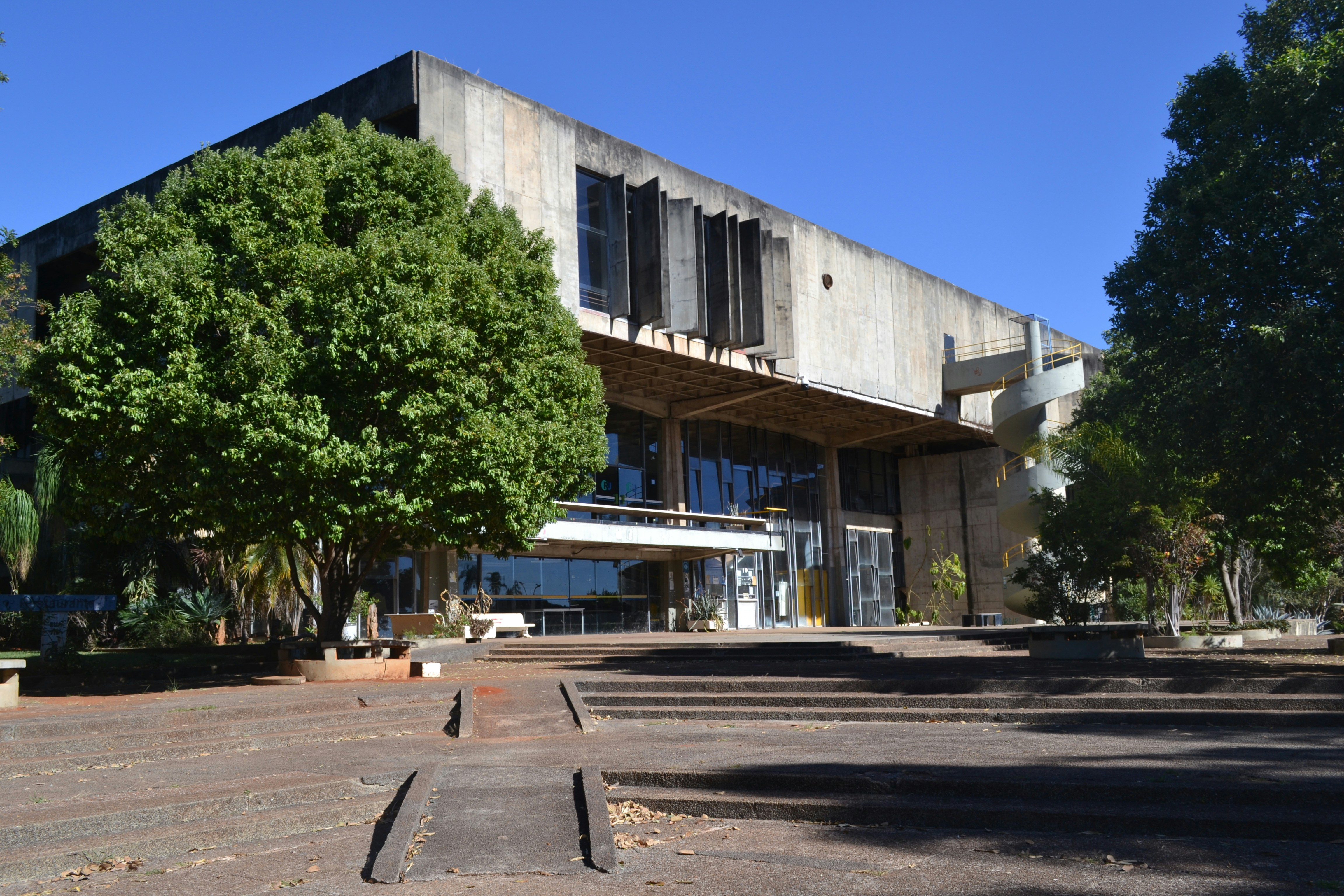 Concrete building with large windows surrounded by lush green trees under a clear blue sky.