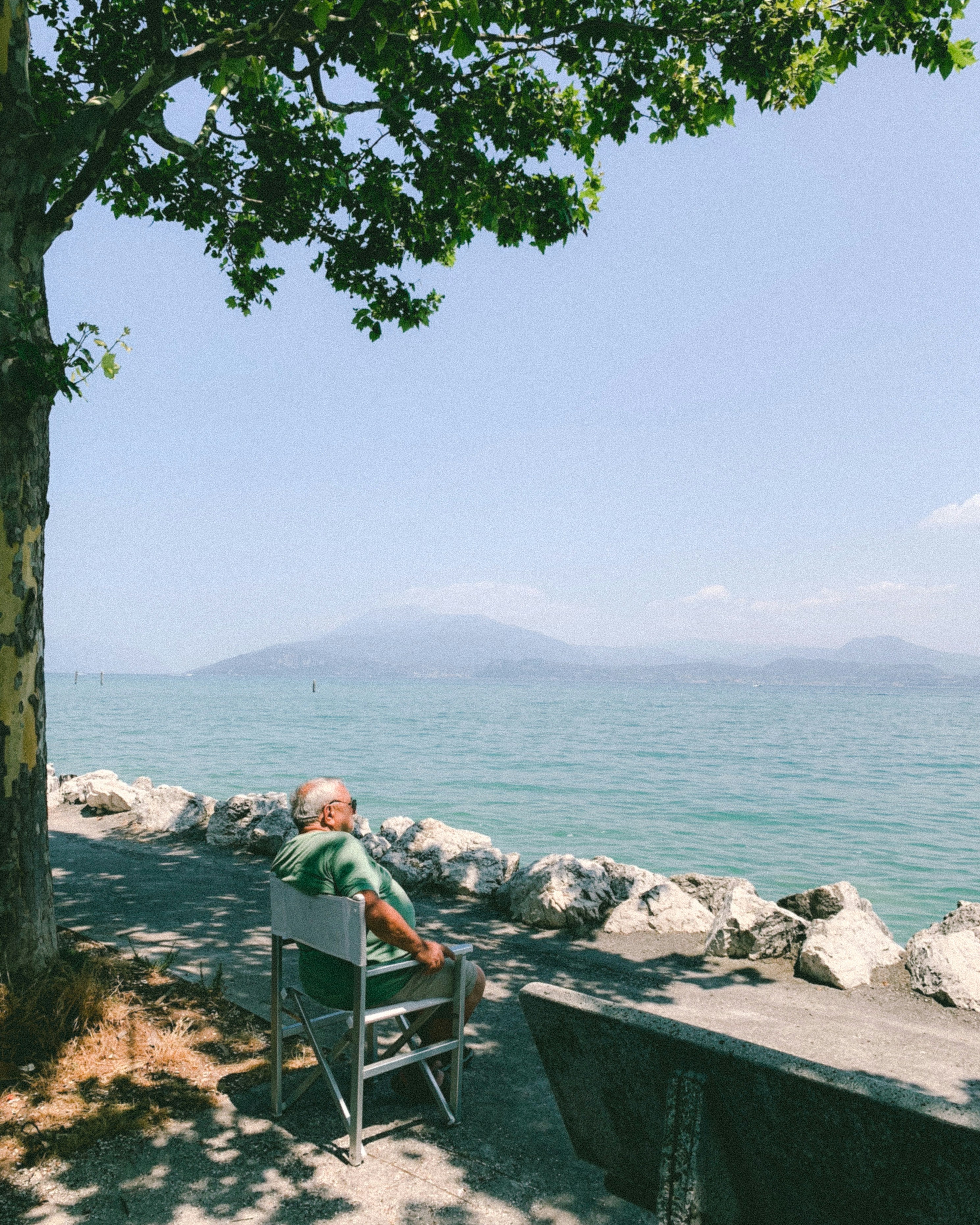 A man relaxes on a chair near the water's edge, framed by a leafy tree, with distant mountains on the horizon.