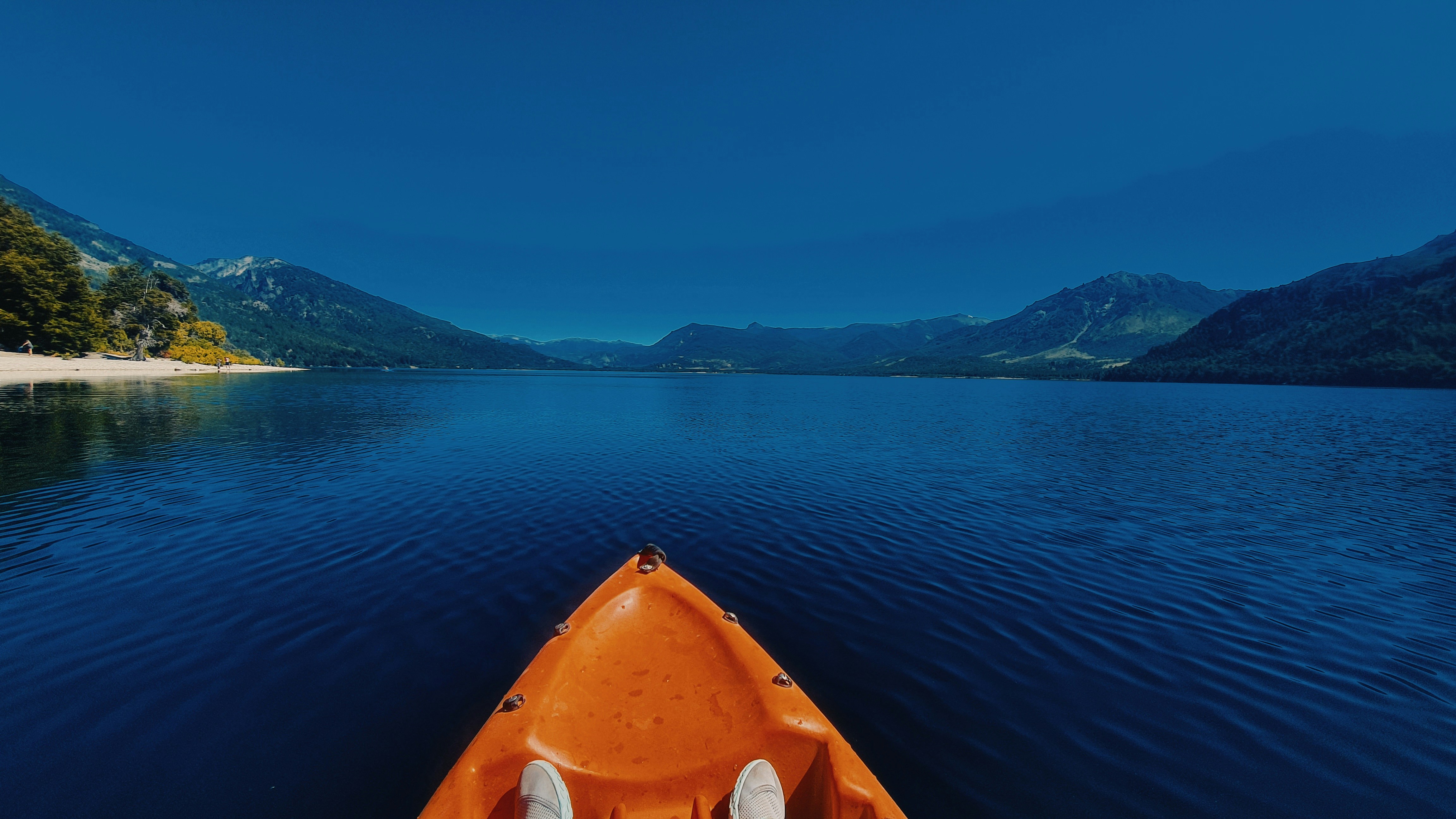 Orange kayak slicing through a calm blue lake surrounded by distant mountains under a clear sky.