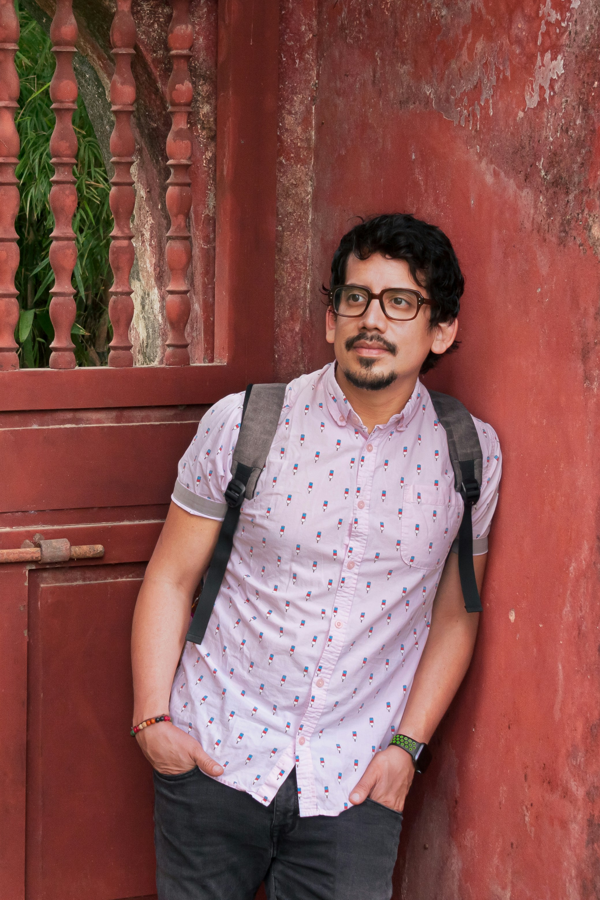 Man in a patterned shirt leaning against a vibrant red wall with a wooden door, surrounded by greenery.
