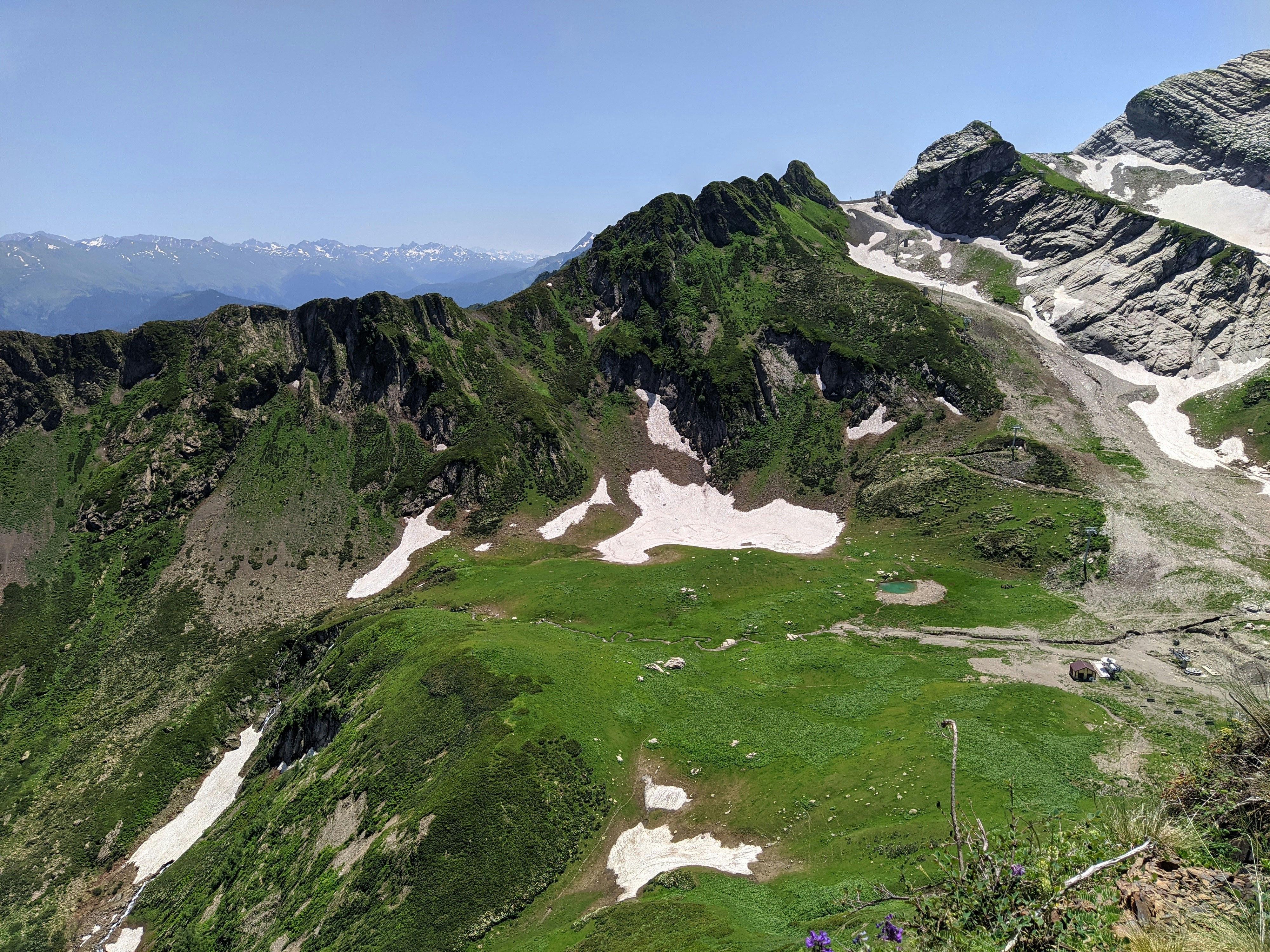 Lush green alpine landscape interspersed with patches of snow beneath a clear blue sky.