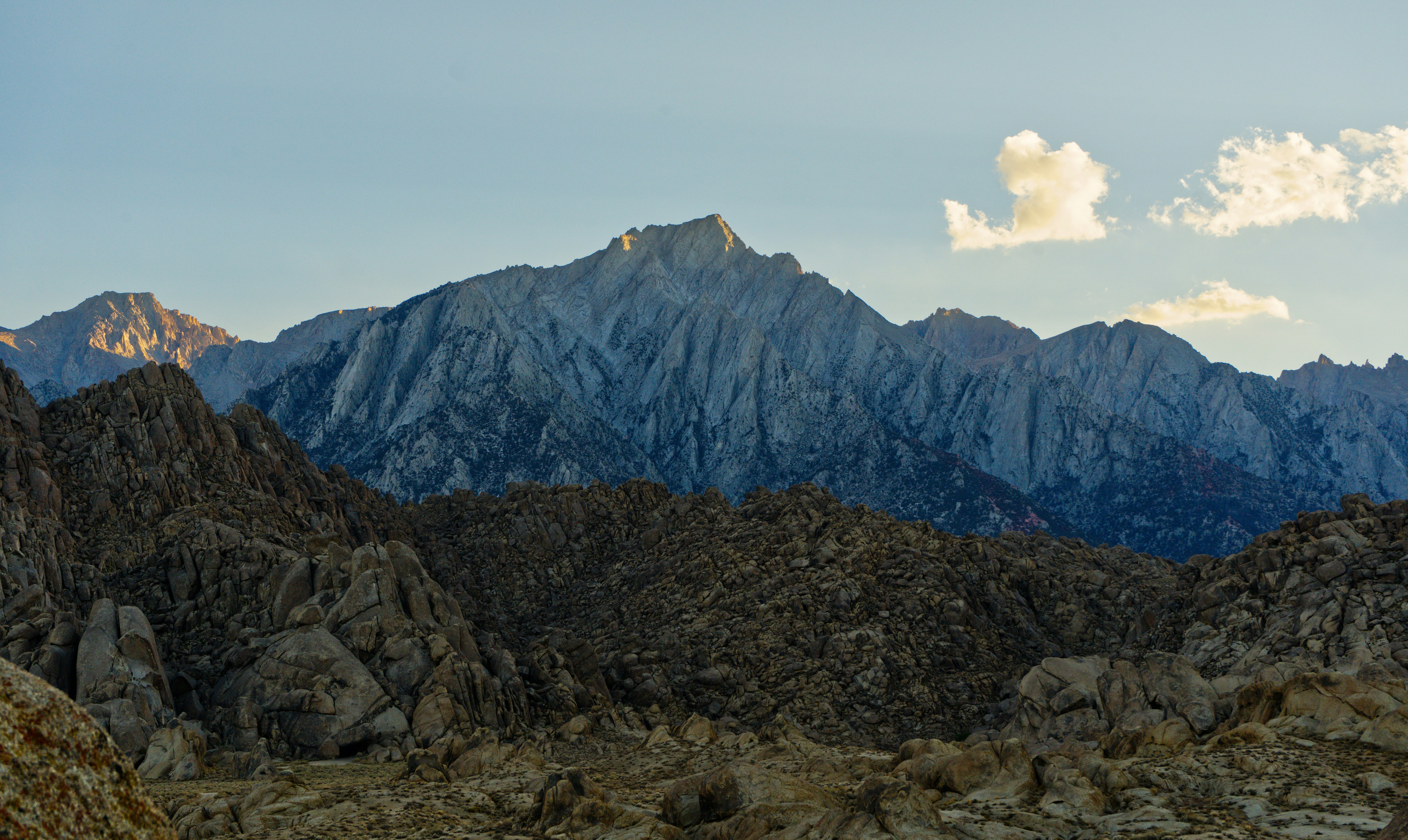 Dramatic mountain range under soft evening light, showcasing rugged peaks and textured rock formations. Clouds drift above, adding depth to the serene landscape.