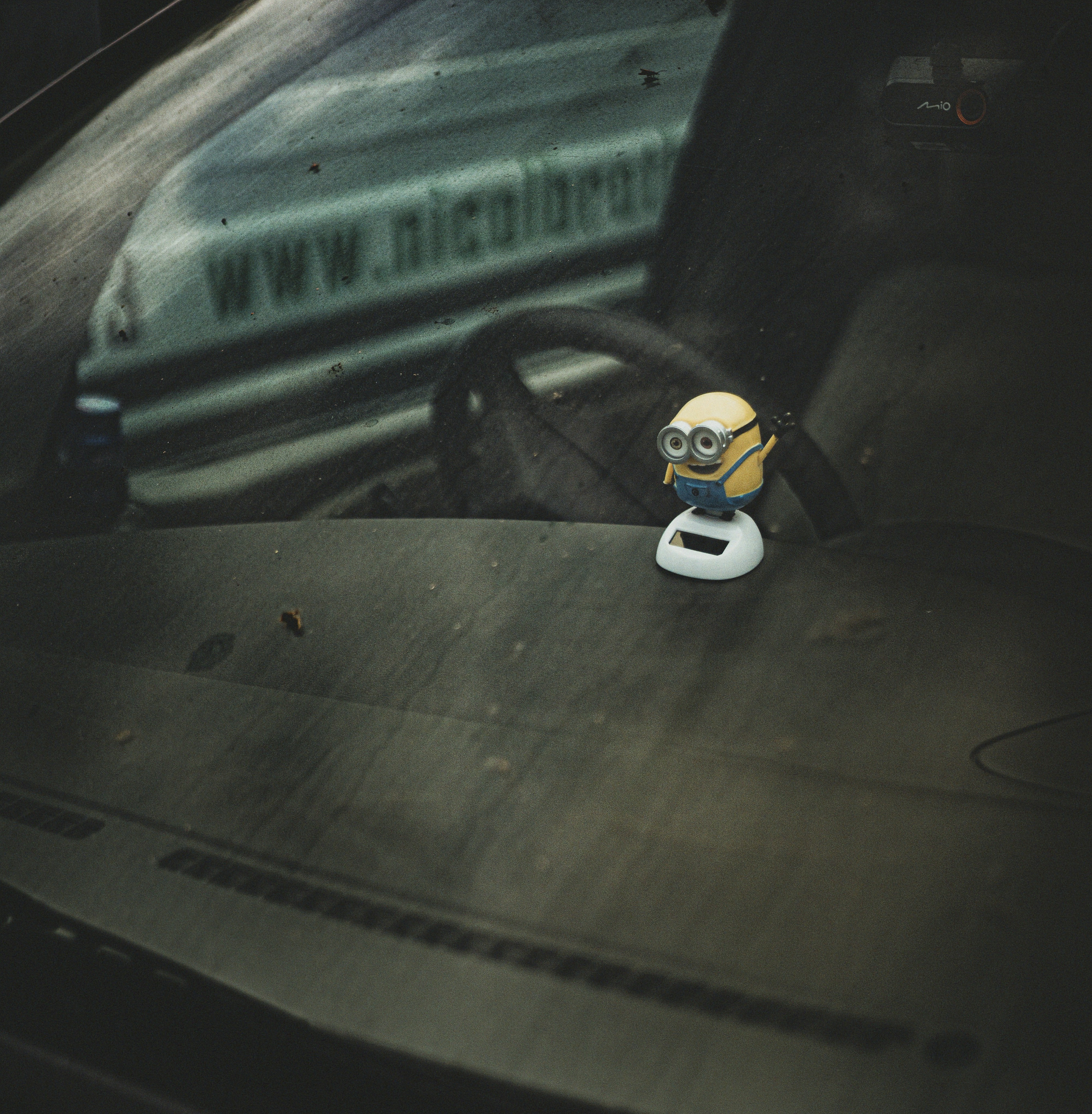 A playful Minion figurine perched on a dusty car dashboard, contrasting with the grime and hints of the outside world.