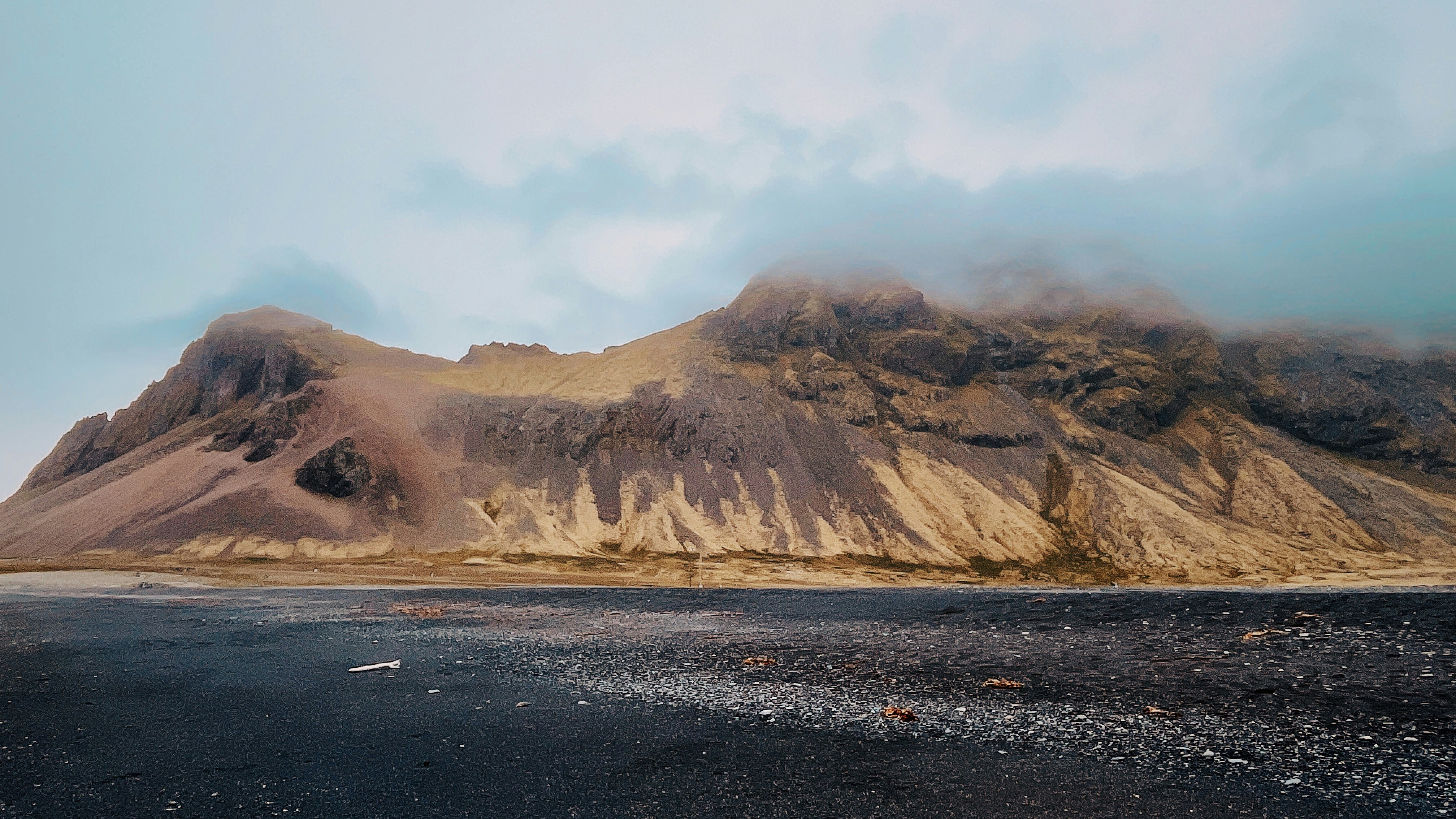 Dramatic mountain landscape shrouded in mist, showcasing layered geological formations and a stark contrast between dark volcanic rock and lighter earth tones.