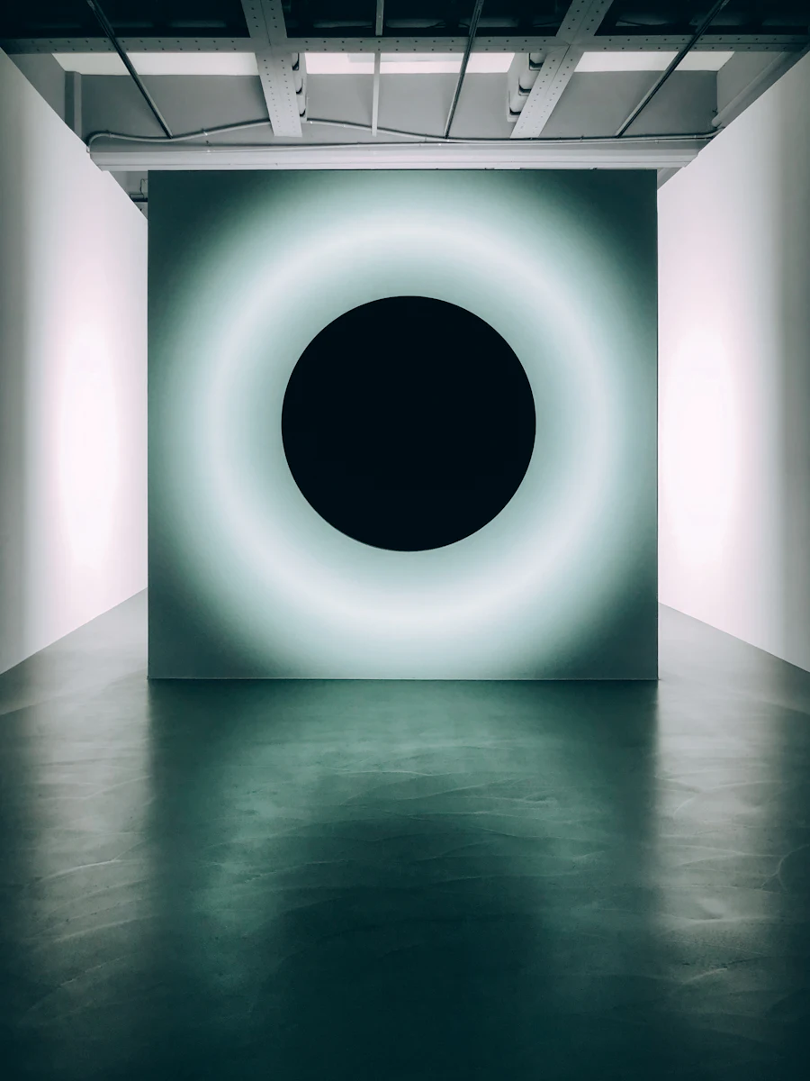 A large square installation with a dark central circle and soft glowing gradient sits in a white gallery space. The polished floor and exposed ceiling frame the artwork symmetrically.