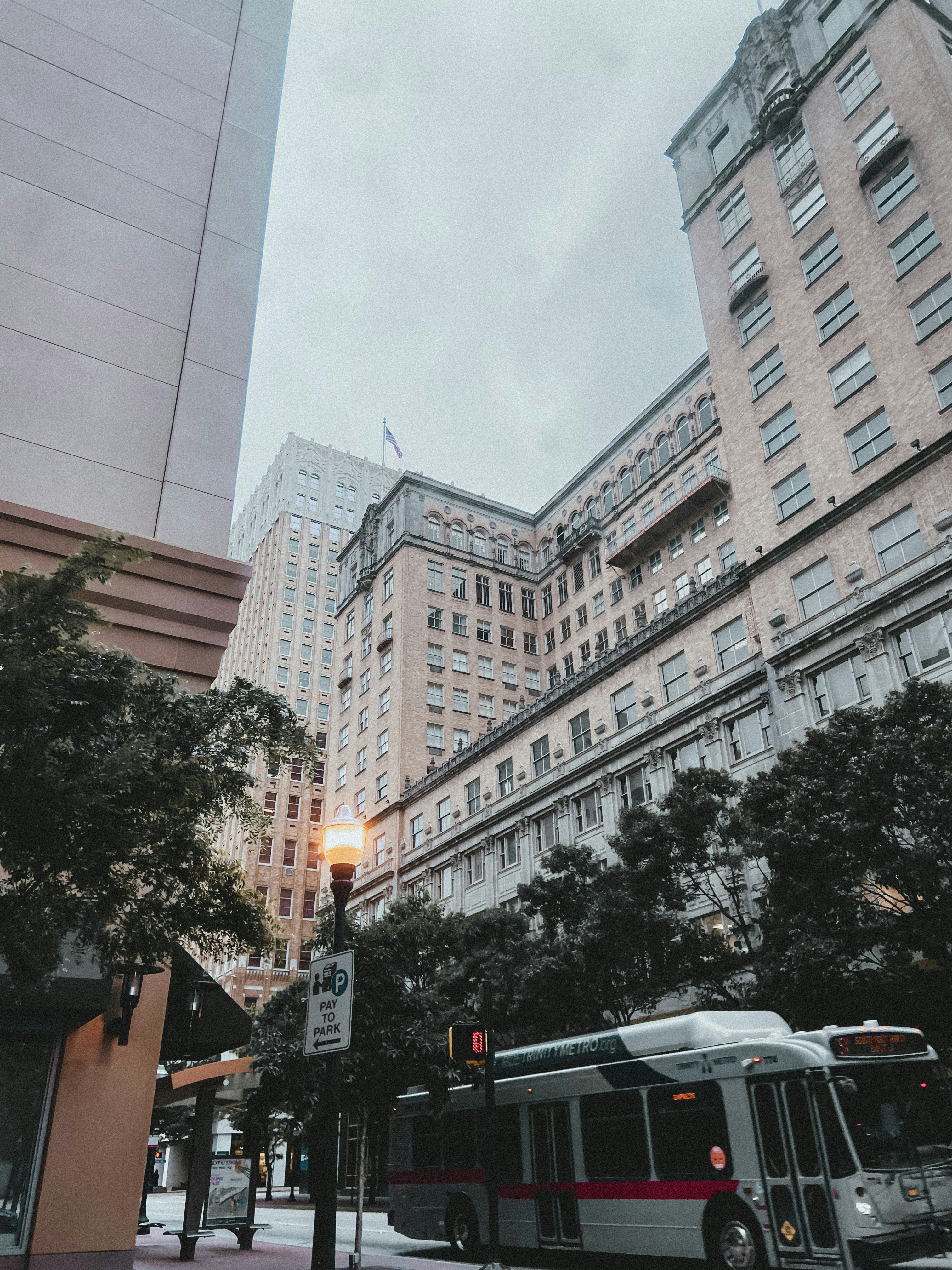 View of towering buildings intersecting with urban foliage, showcasing a blend of historical and modern architecture. A city bus navigates through the scene.