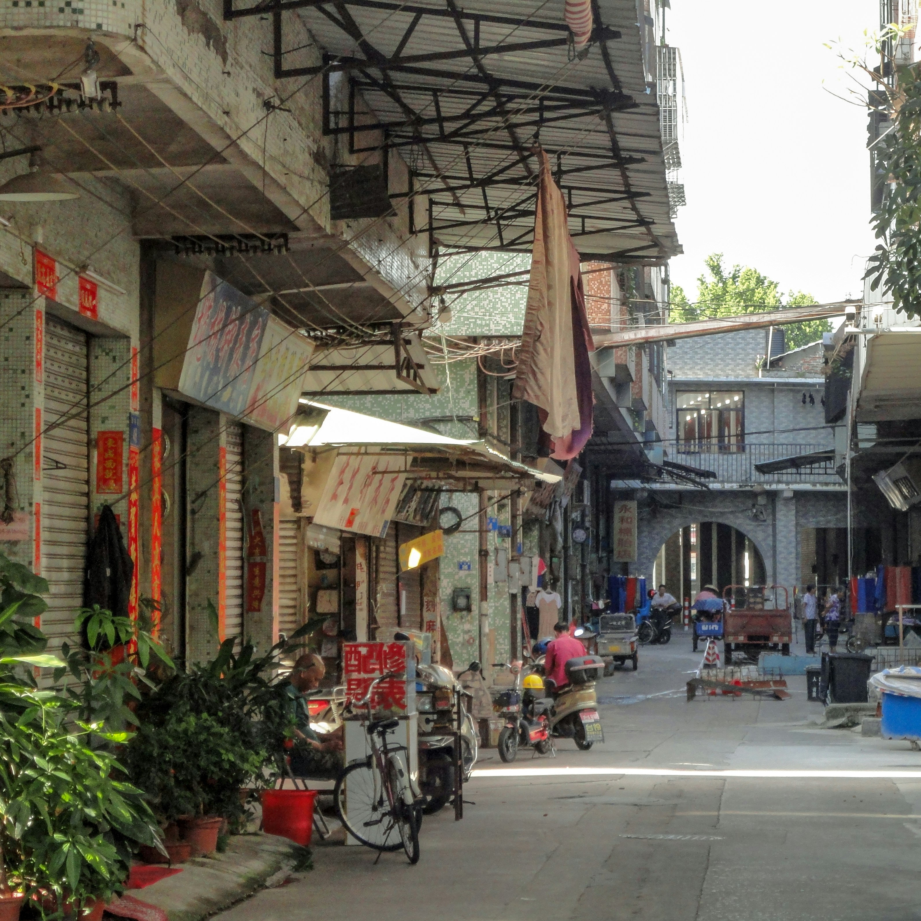 Narrow alleyway lined with shops and plants, leading to an archway that hints at hidden stories. A cyclist passes through, adding life to the scene.