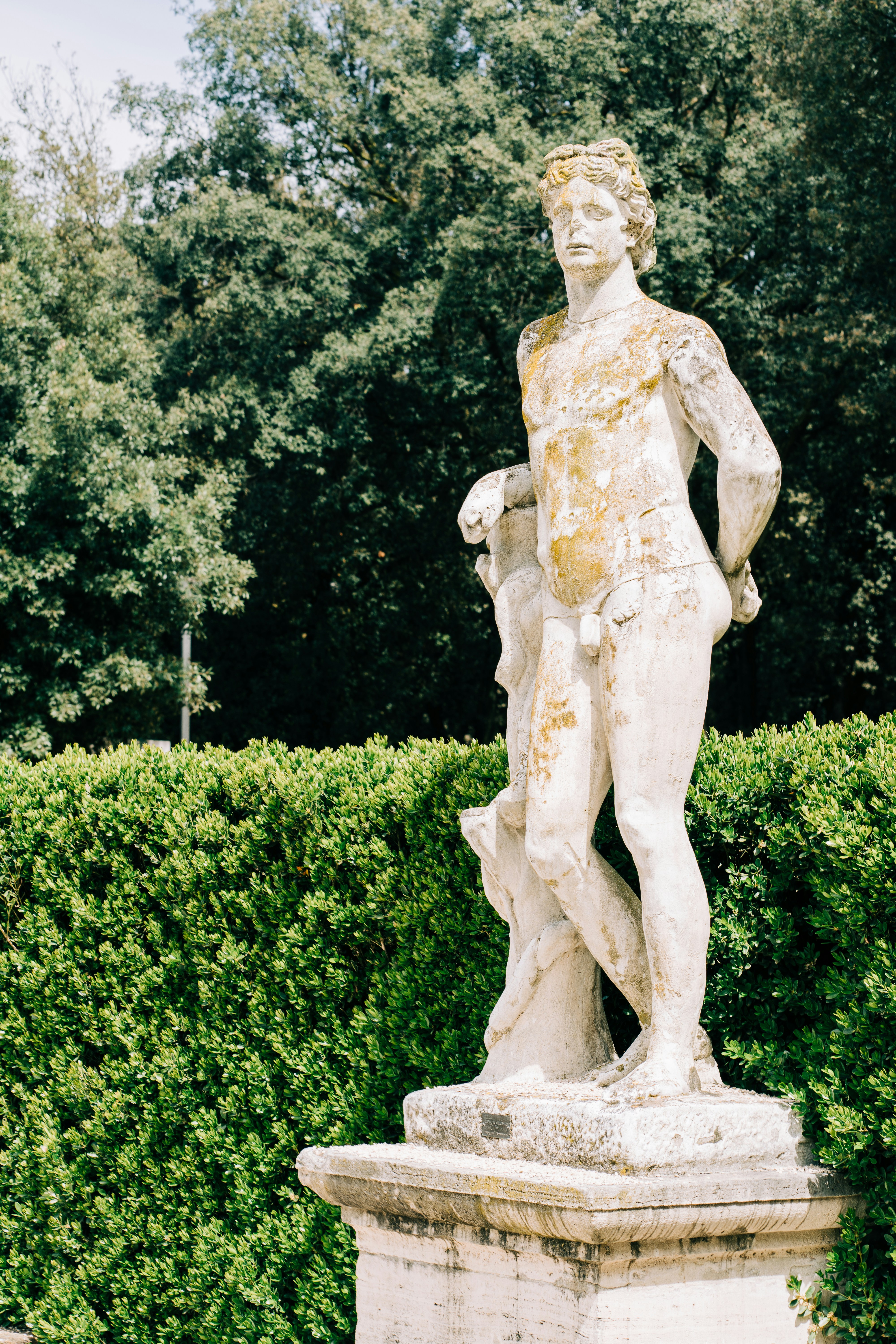 Weathered marble statue depicting a male figure in a dynamic pose, surrounded by lush greenery.
