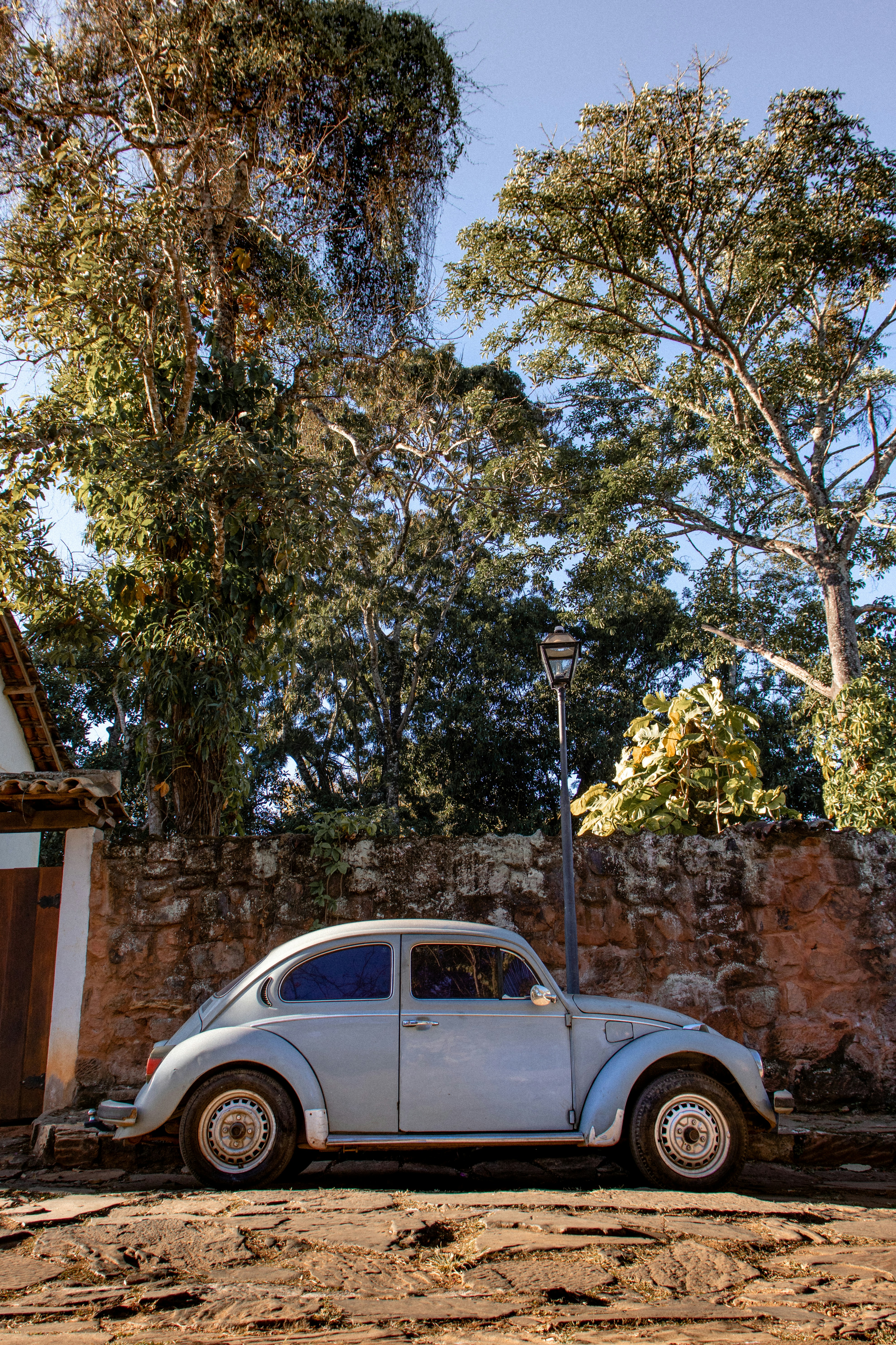 Classic blue Volkswagen Beetle parked beside a textured stone wall, framed by lush greenery and a vintage street lamp.