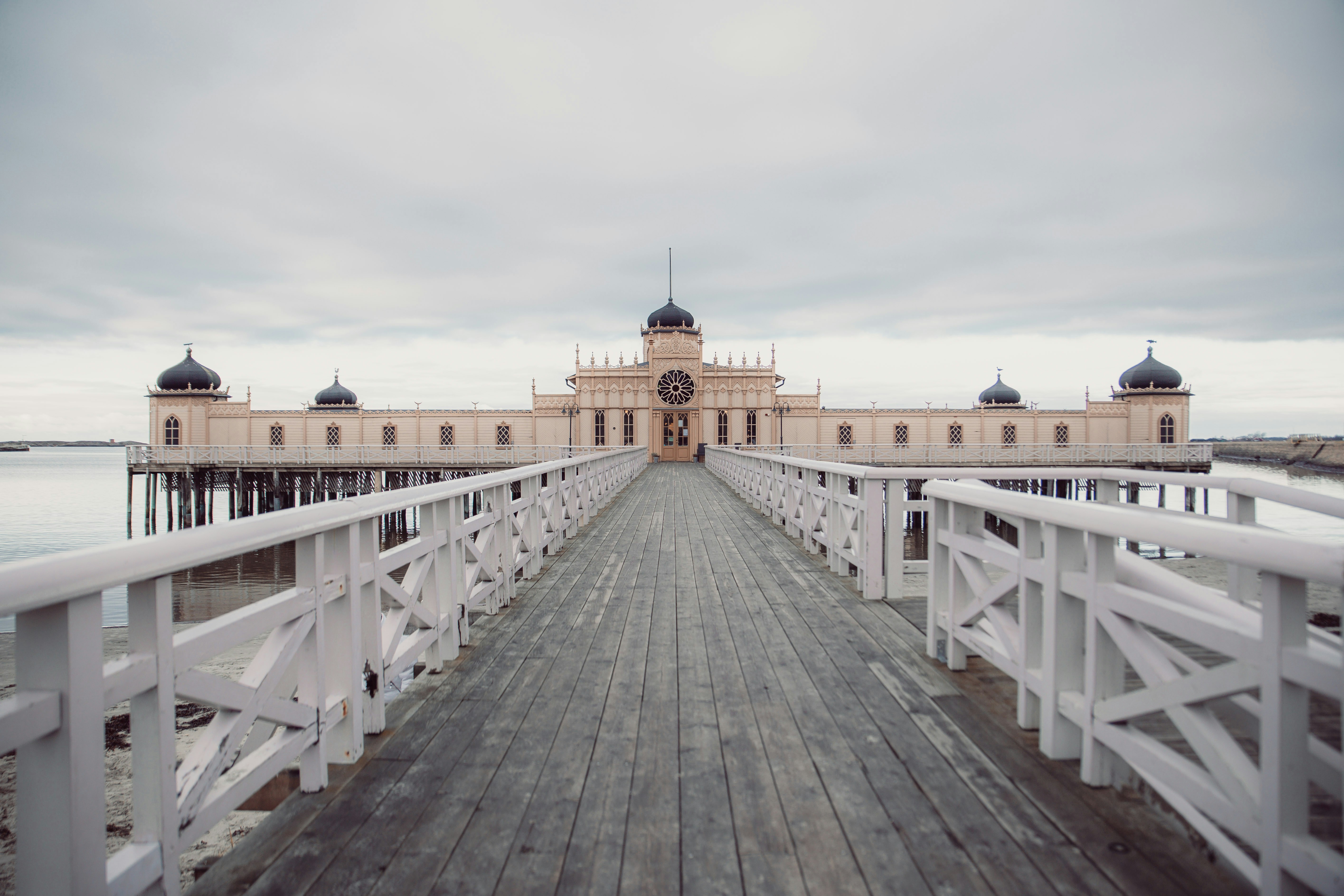 Historic pier extending towards a grand seaside building with intricate architectural details under a cloudy sky.