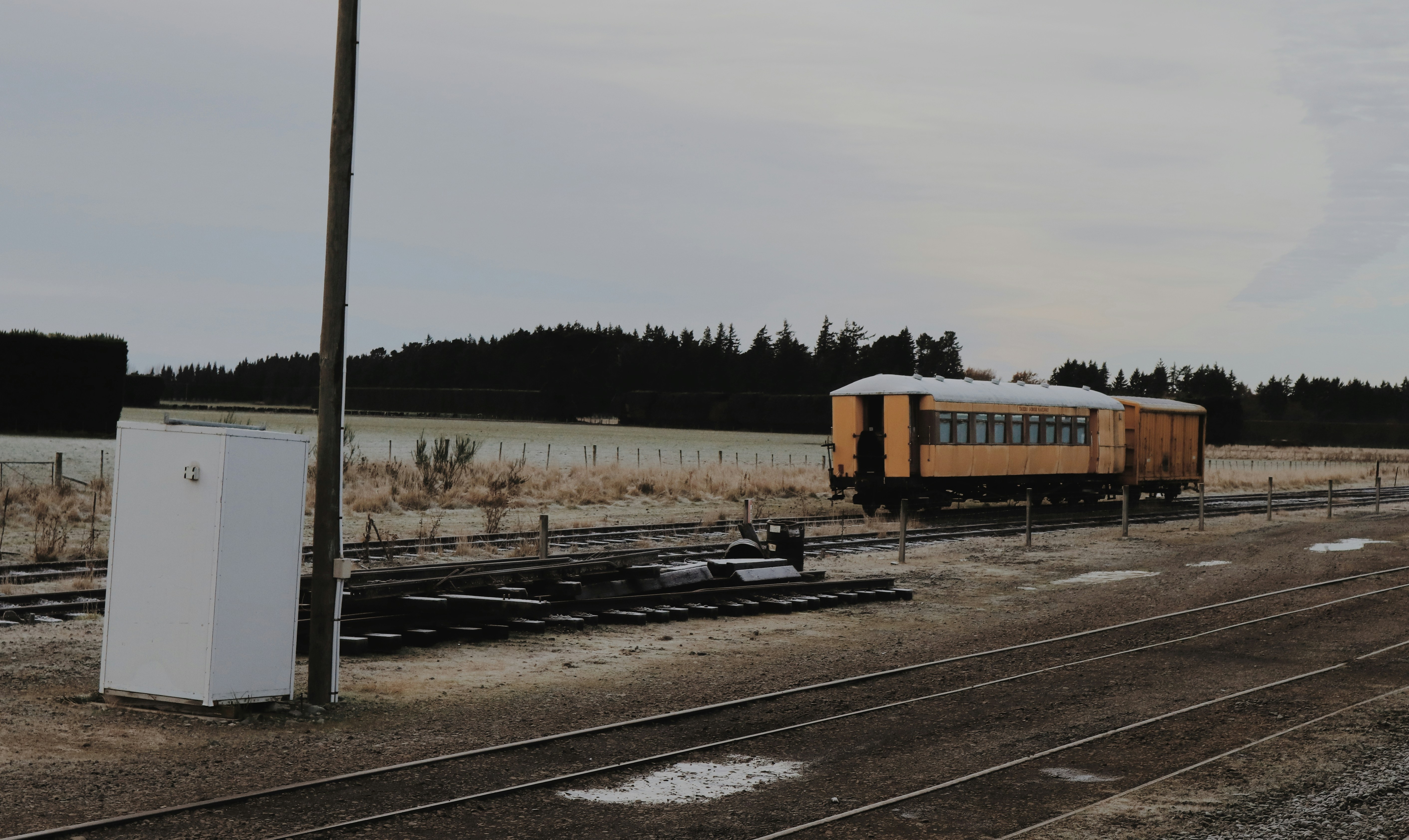 Abandoned wooden train carriage resting on quiet tracks, surrounded by a tranquil landscape of grass and distant trees.