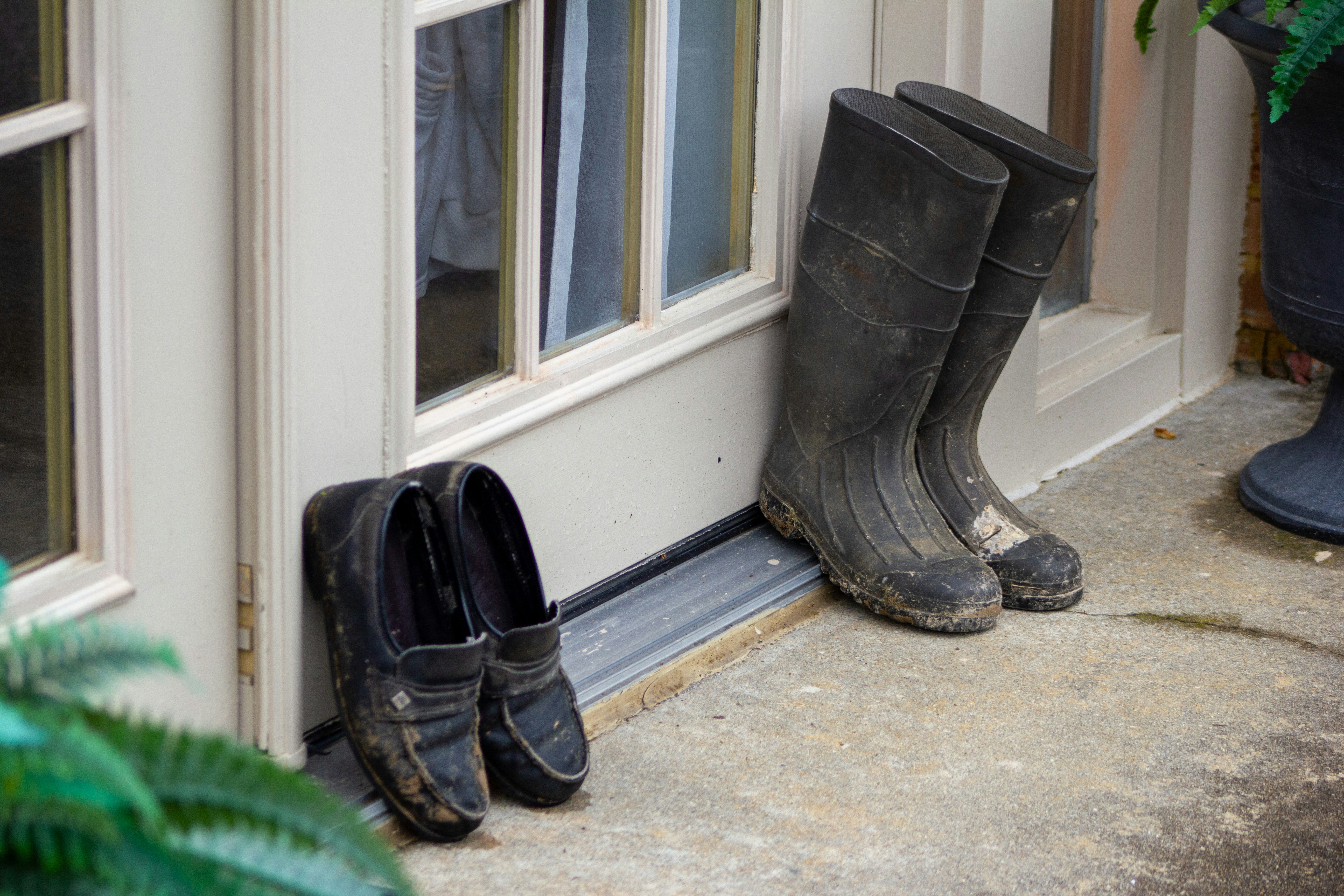 Black leather boots and muddy rubber boots next to a white framed glass door.
