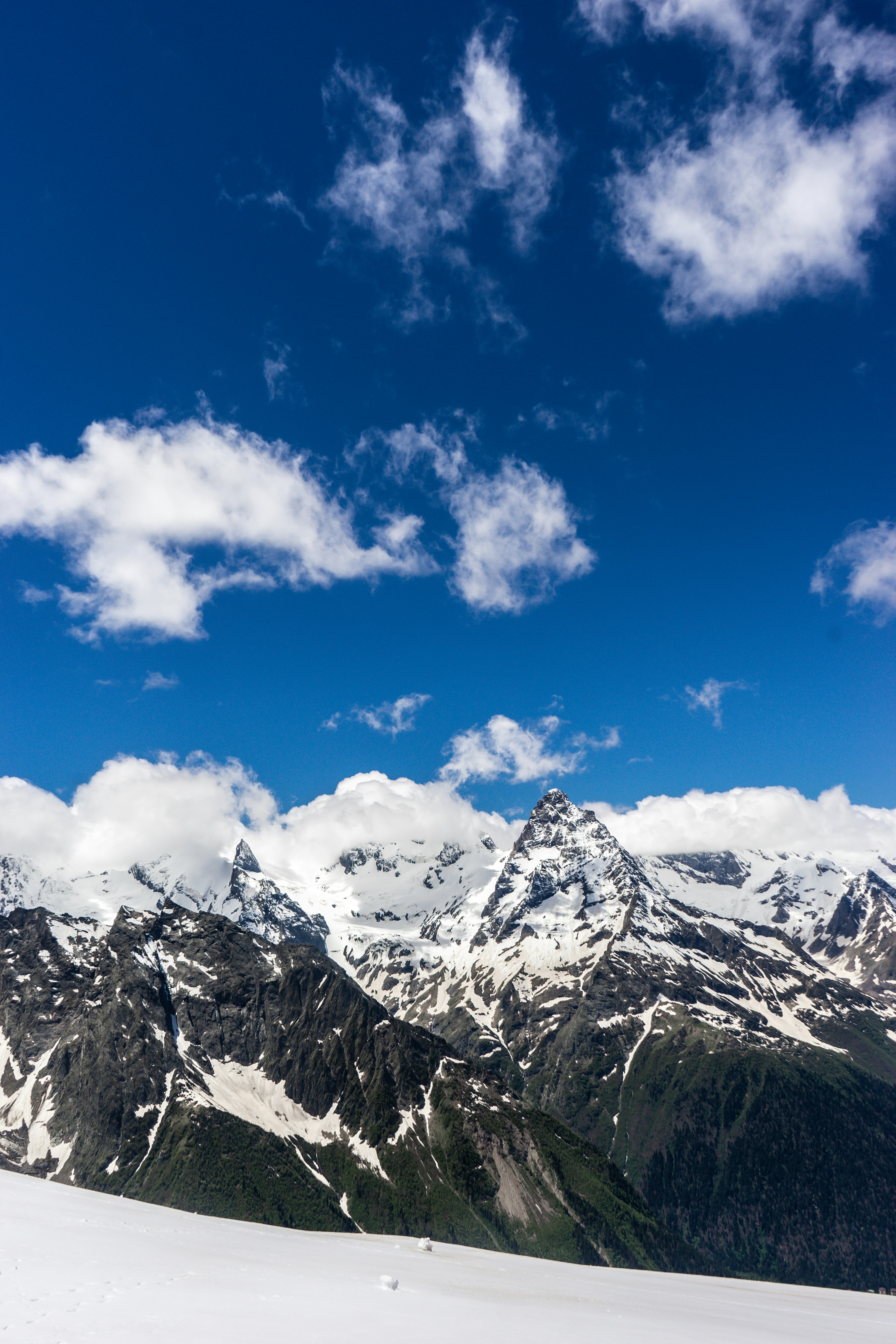 Snow-capped mountain range stretches across the horizon beneath a vibrant blue sky dotted with fluffy clouds.