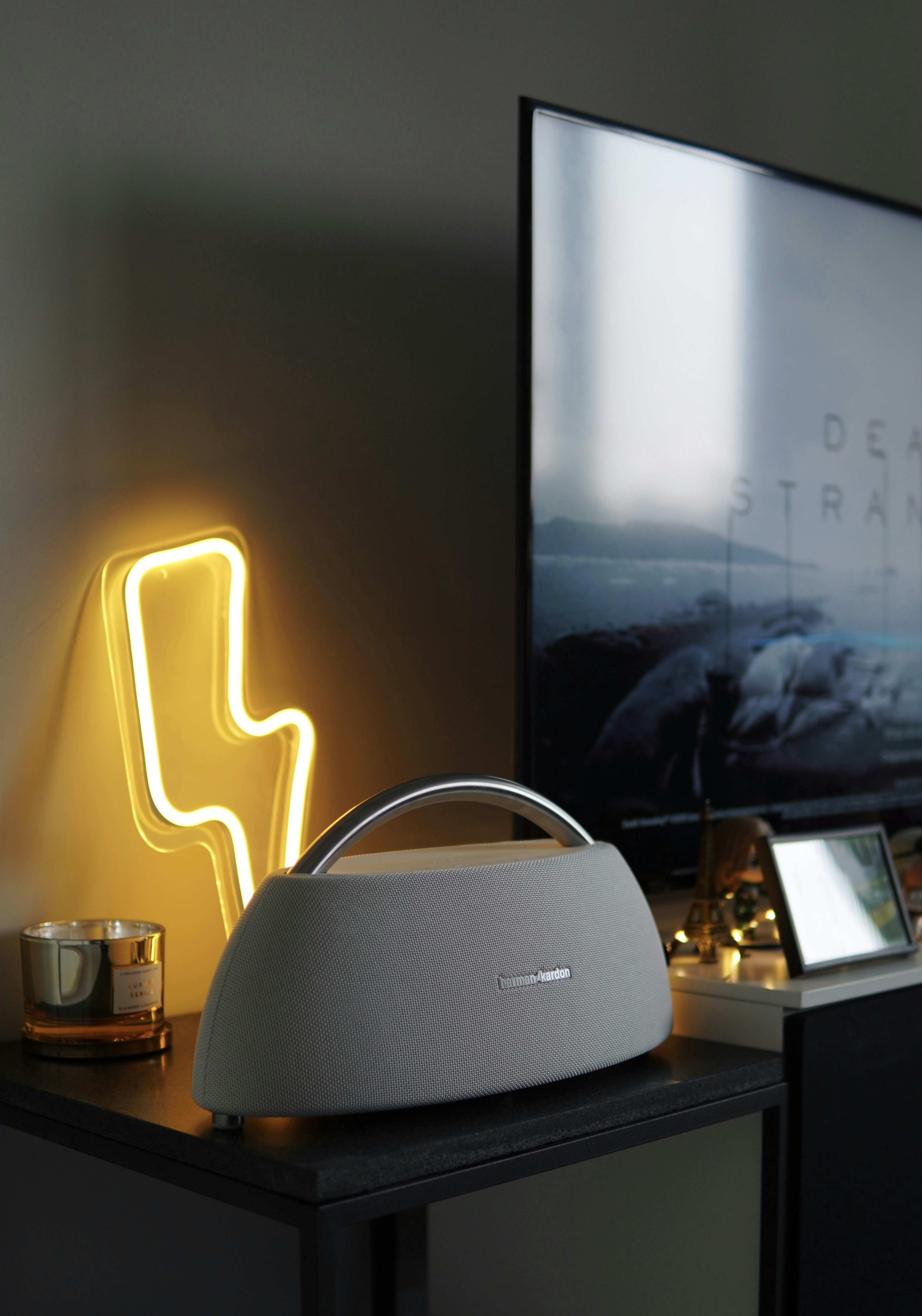 Gray wireless speaker sits on a dark desk beside a neon-yellow lightning bolt sign. A large screen in the background shows a moody landscape.