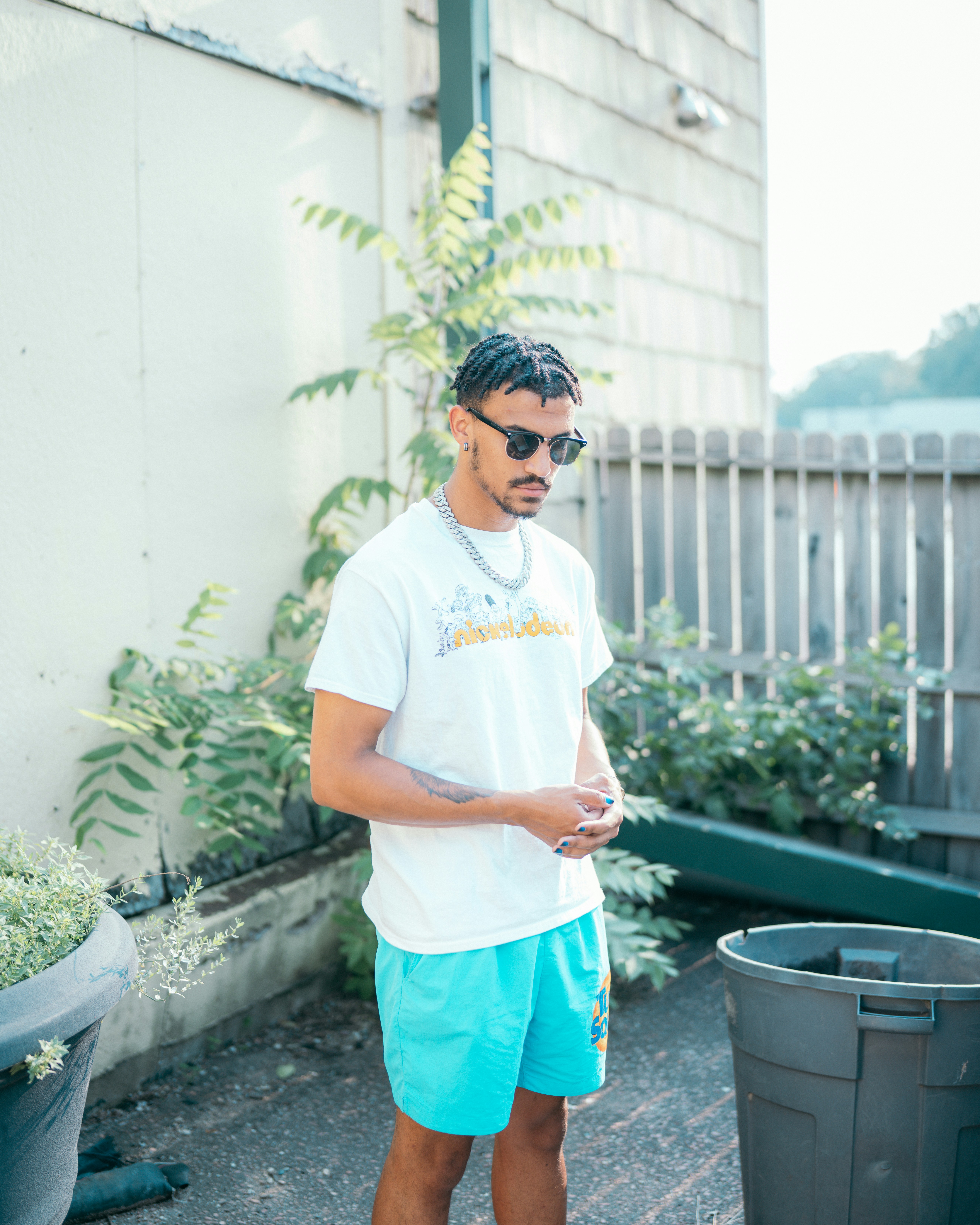 Young man in casual attire stands thoughtfully beside a planter in an urban setting, with greenery in the background. 