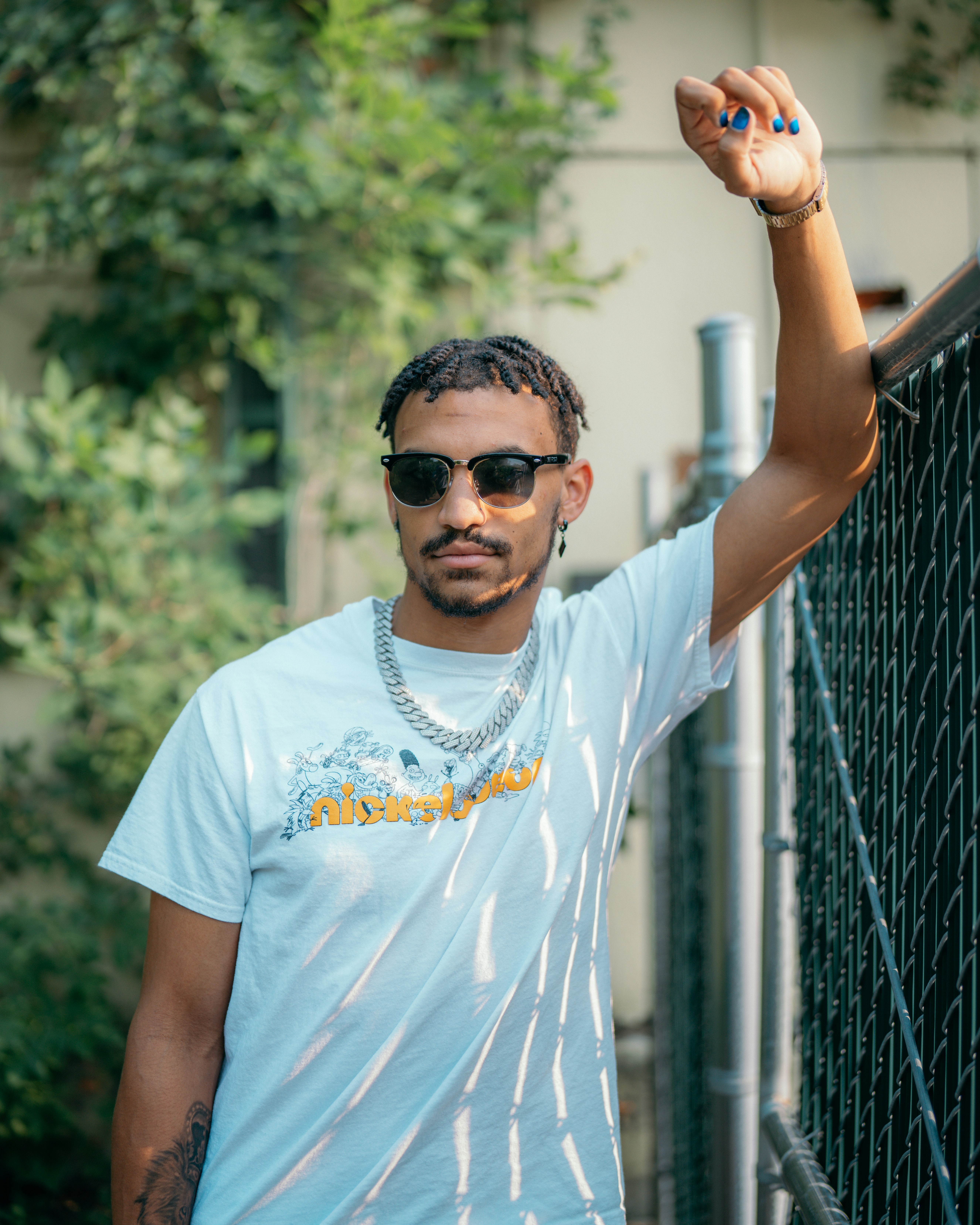 Young man leaning against a fence, wearing sunglasses and a casual t-shirt, surrounded by greenery.