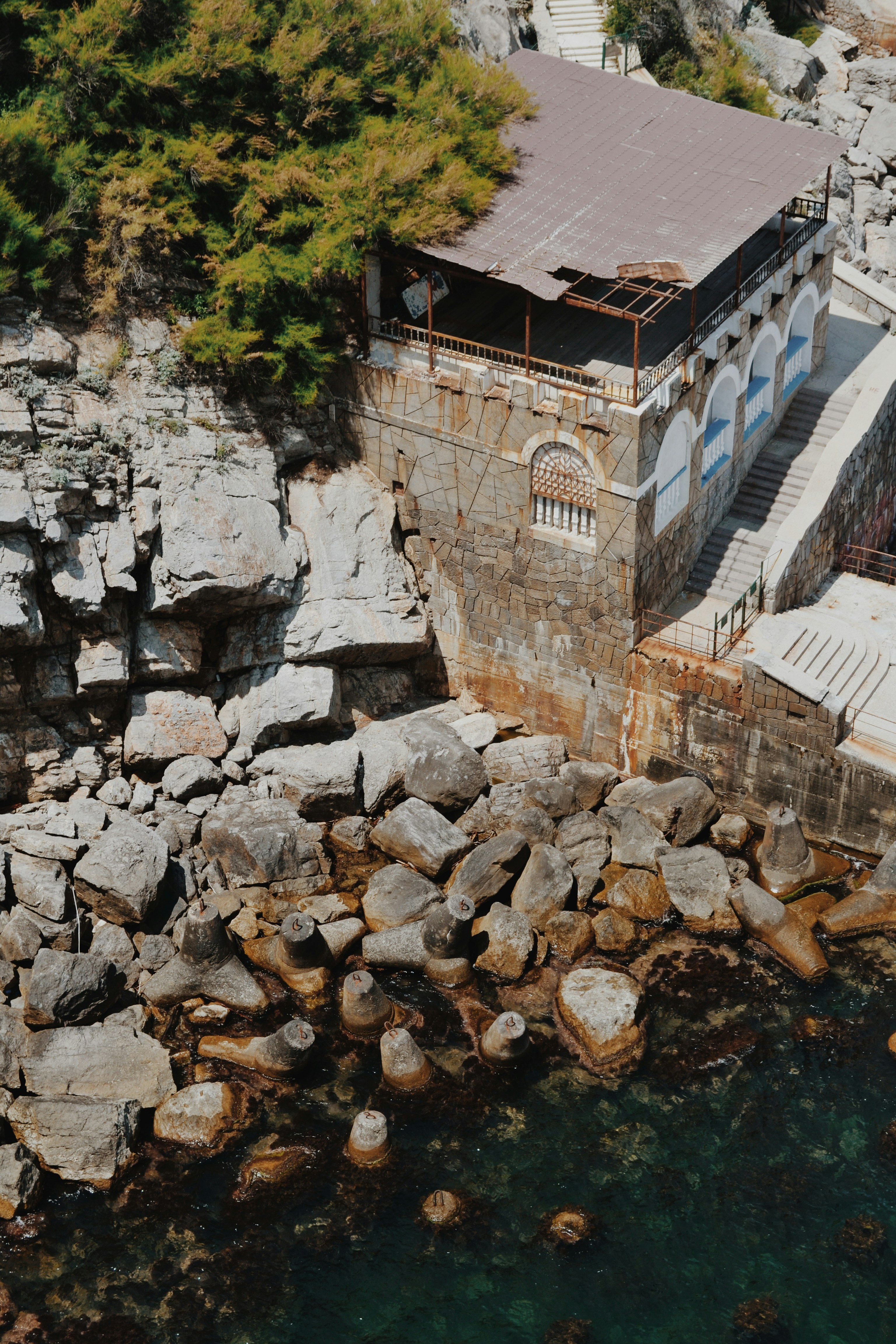Rustic stone building perched on rocky coastline, surrounded by lush greenery and clear waters. Stairs lead down to the shoreline.
