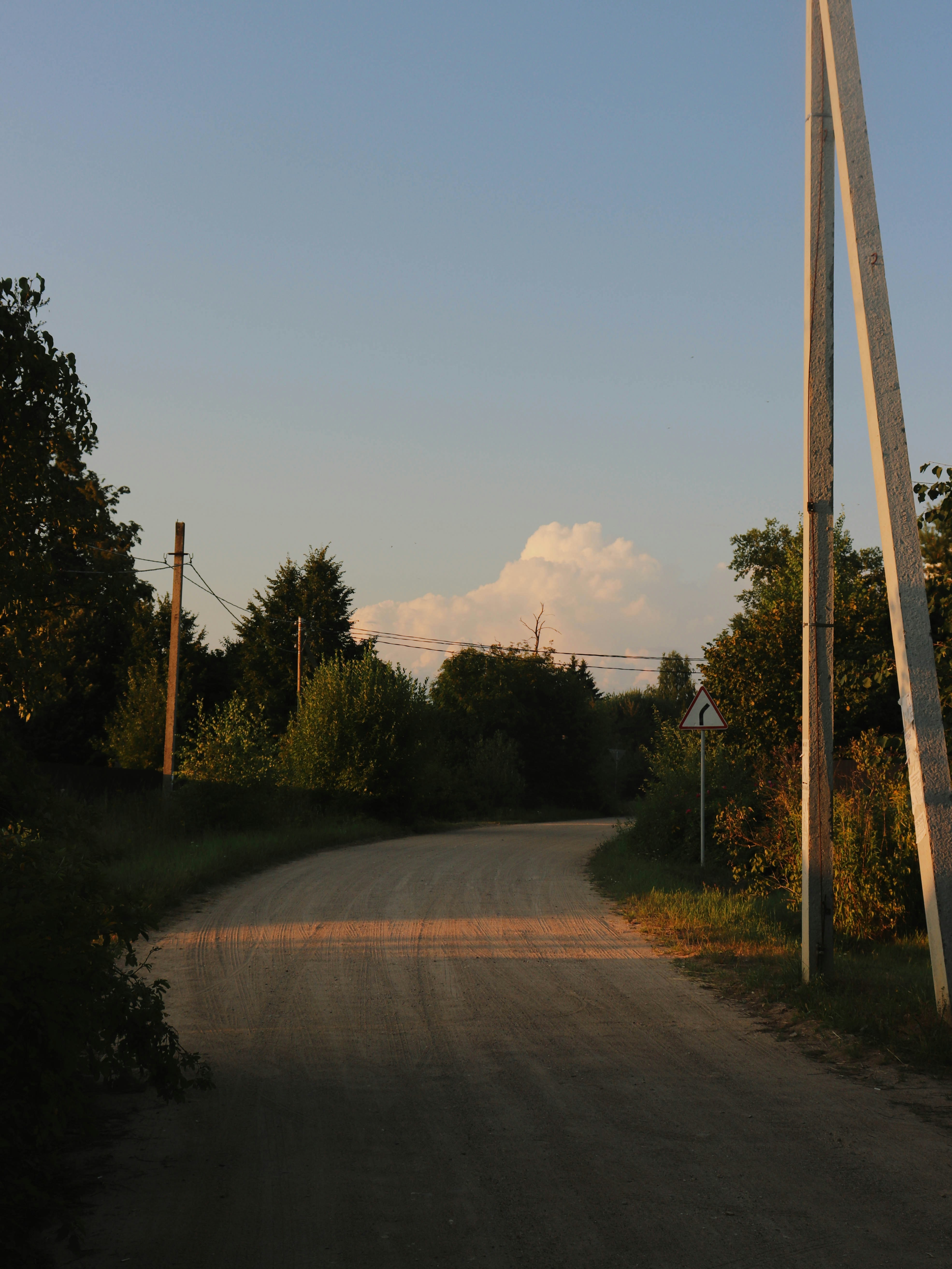 Winding dirt road bordered by lush greenery under a soft evening sky, leading towards distant clouds. A solitary road sign hints at the journey ahead.
