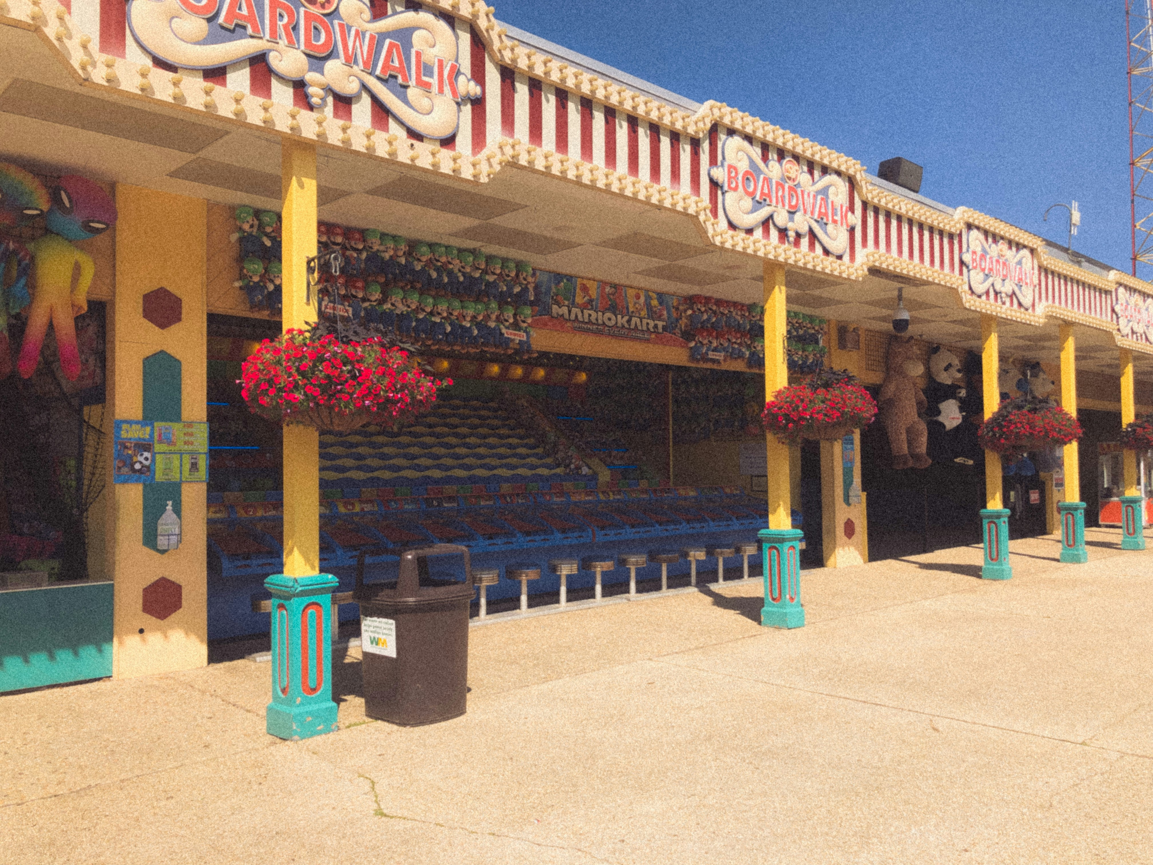Colorful boardwalk game stall adorned with vibrant flowers and plush toys, inviting visitors to try their luck. A nostalgic atmosphere reminiscent of summer fairs.