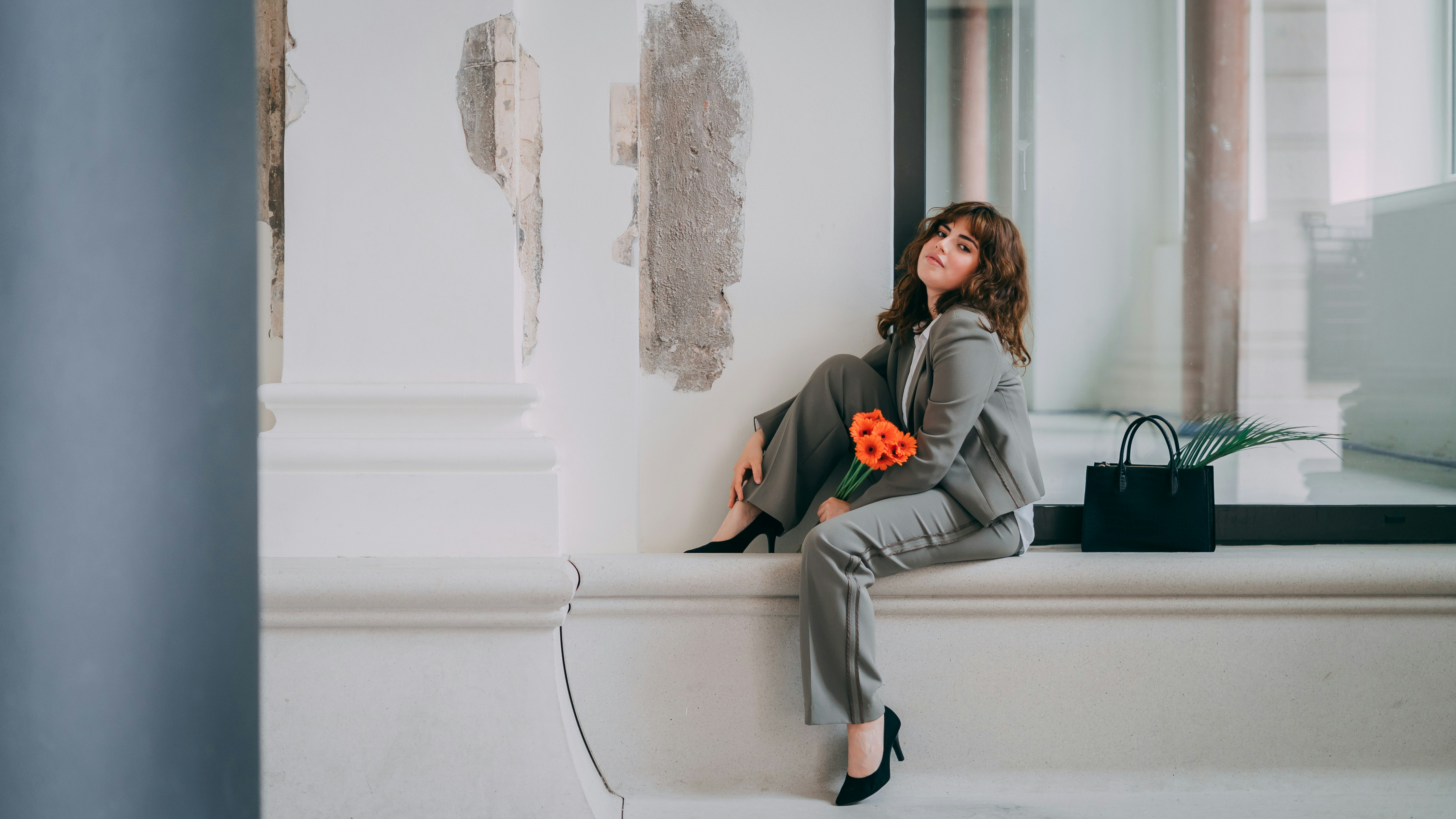 A woman in a tailored gray suit poses gracefully with orange flowers, set against a backdrop of minimalist architecture and soft natural light.