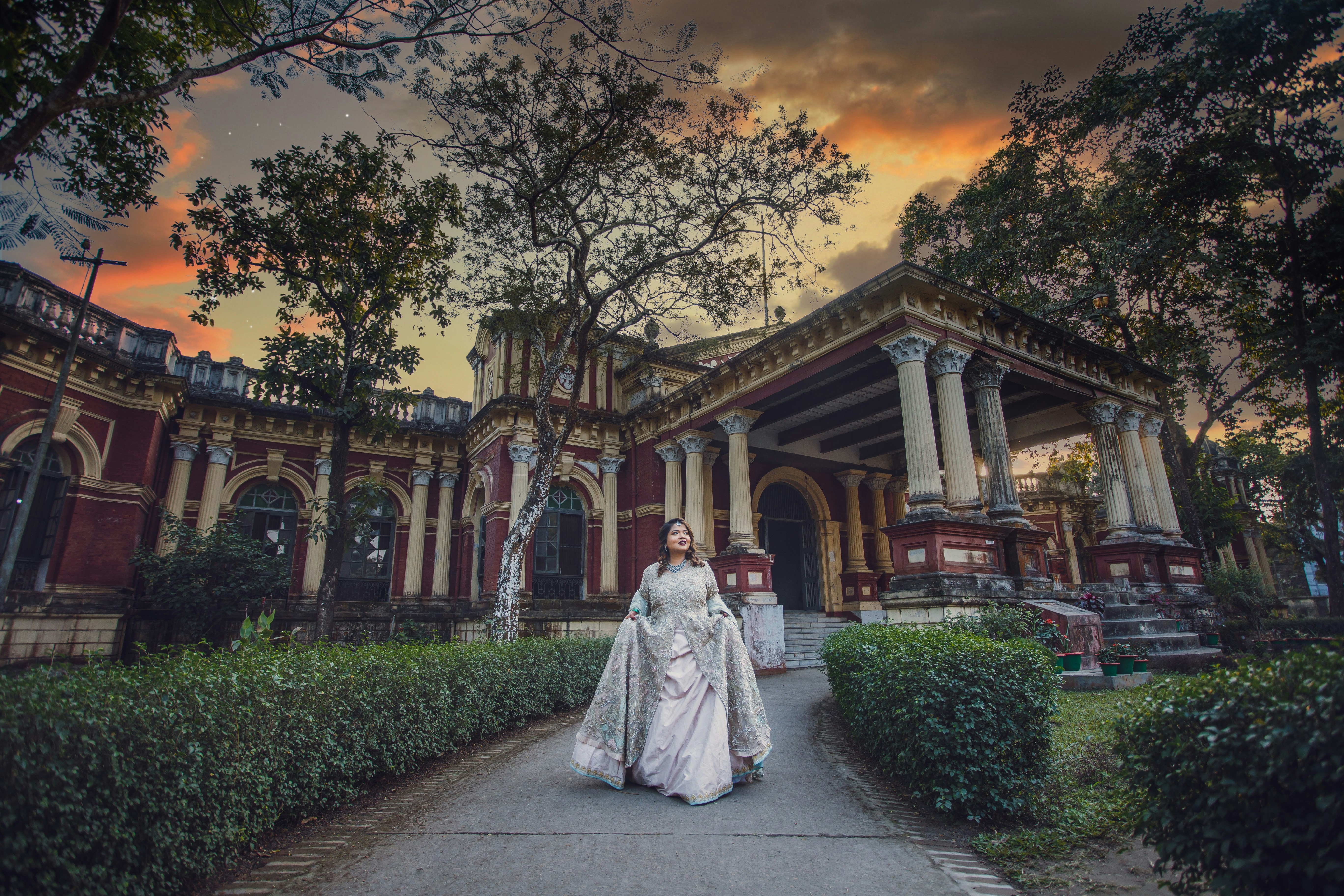 A woman in an ornate gown walks along a pathway framed by lush greenery and a grand, historical building. The scene is illuminated by a dramatic sunset.