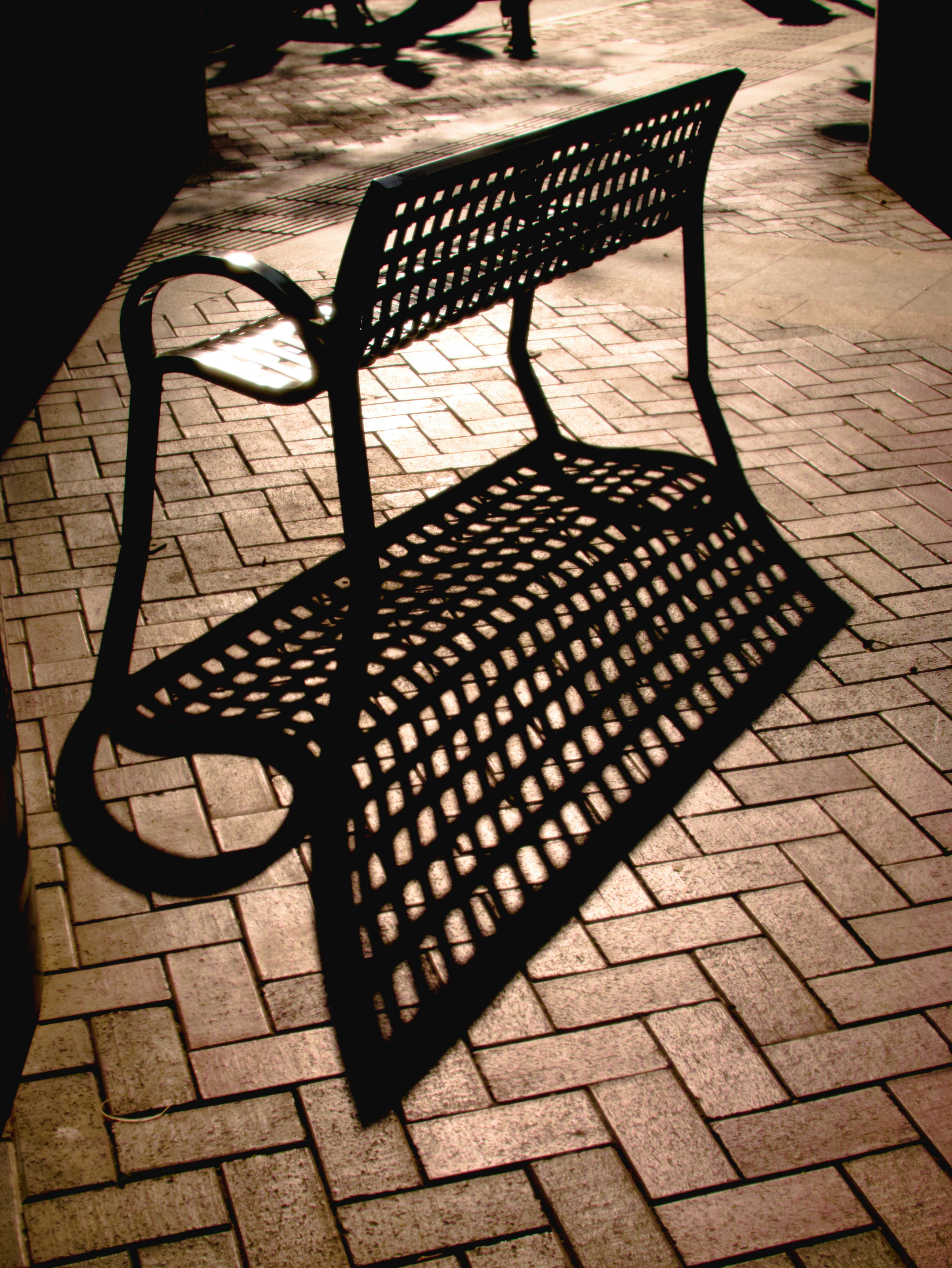 A bench casts intricate shadows on a patterned brick surface, highlighting the play of light and form in an urban setting.