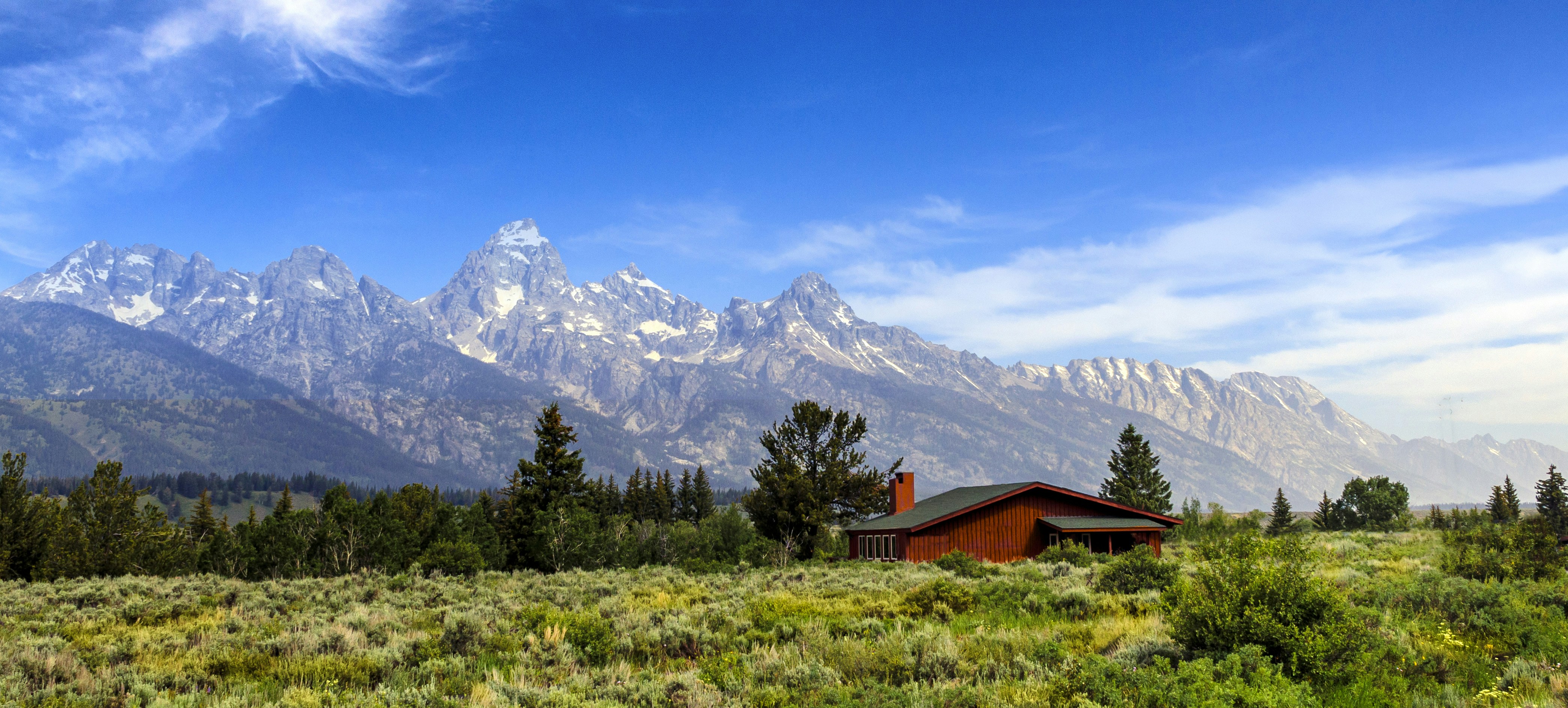A modern cabin nestled among the lush greenery, with majestic mountains rising in the background under a clear blue sky.