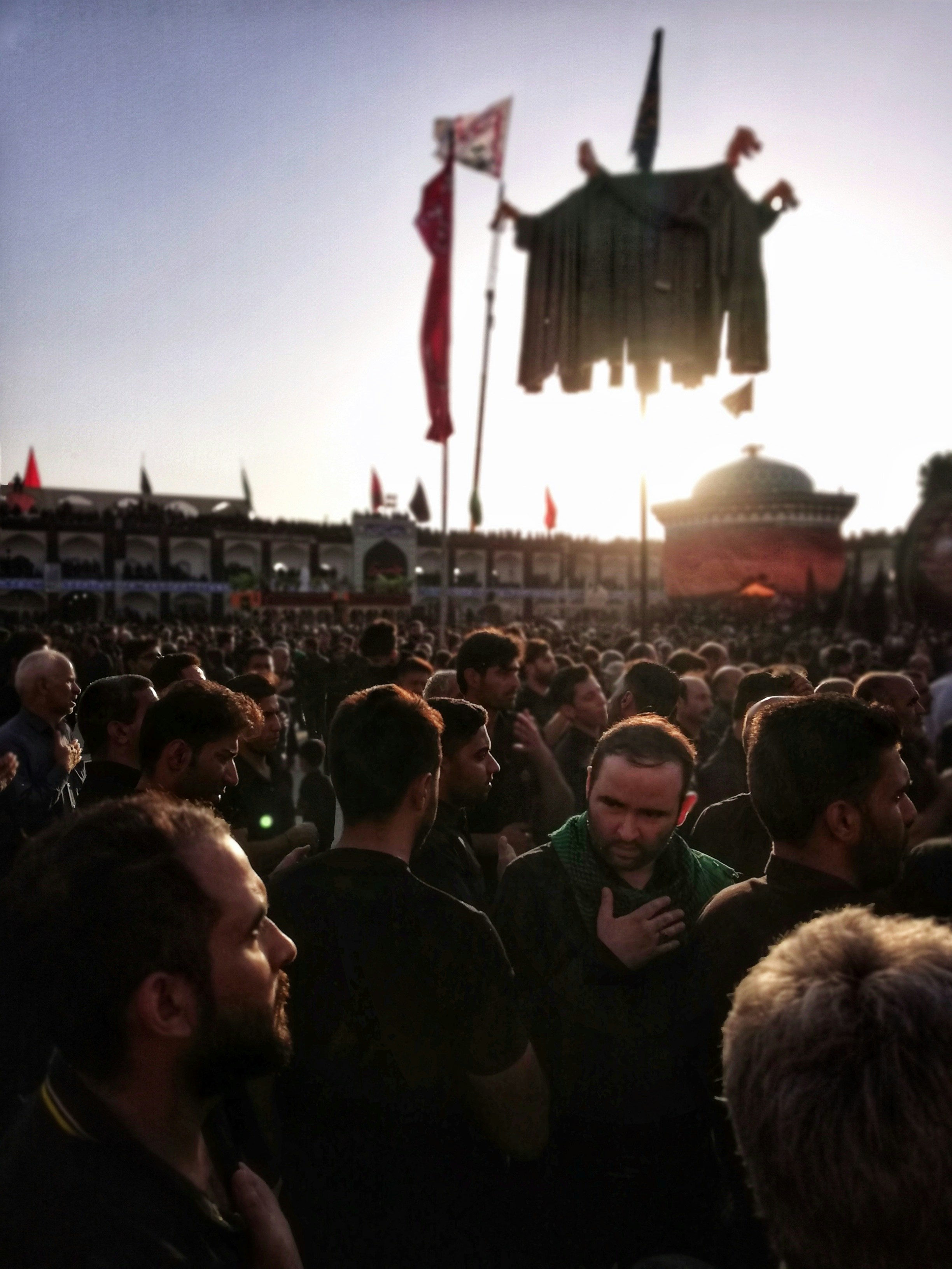 Crowd of mourners gathered during a religious procession under a large flag and canopy at sunset.