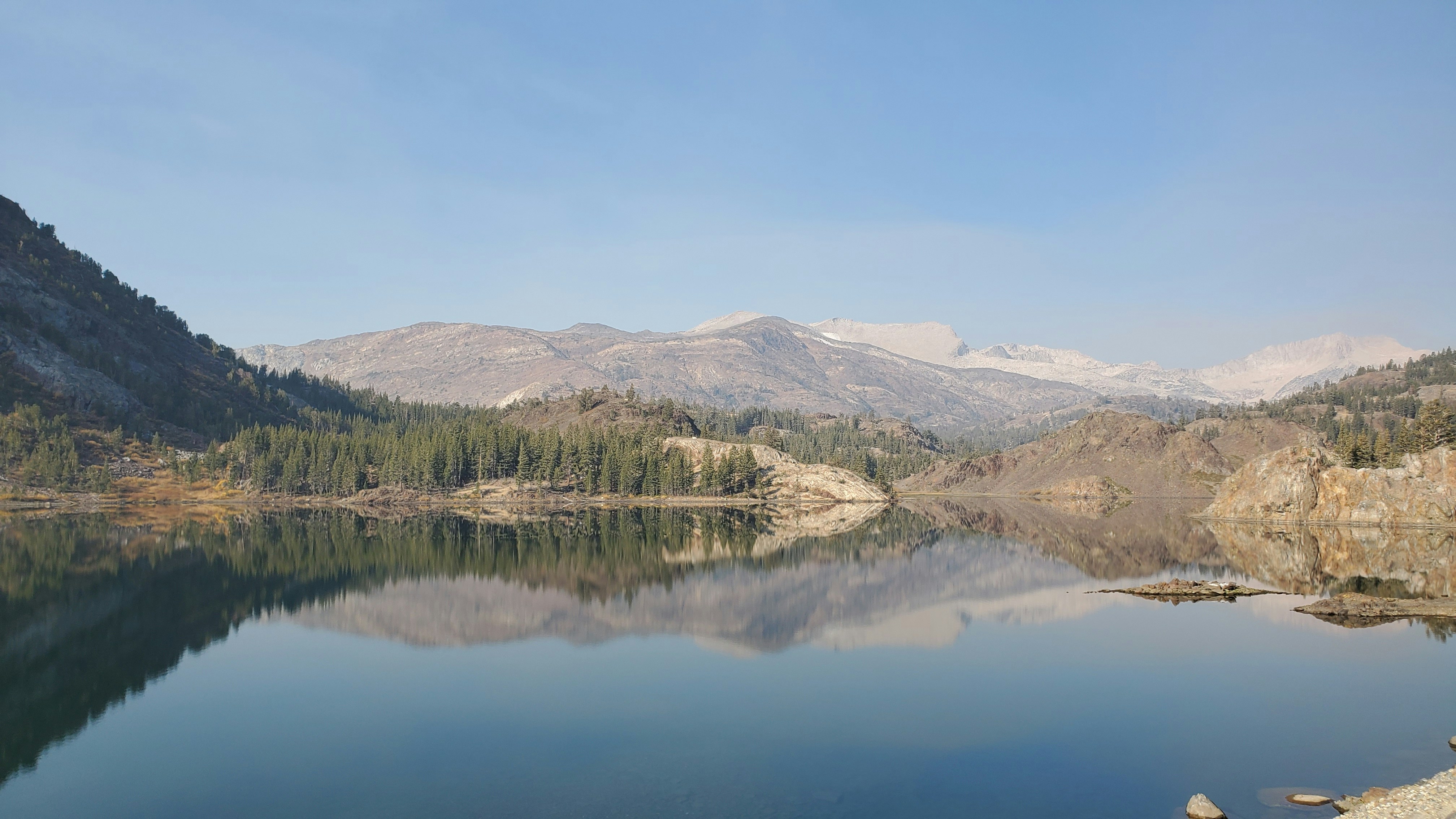 Tranquil mountain lake reflecting surrounding peaks and trees under a clear sky. The still water creates a mirror-like effect, enhancing the natural beauty.