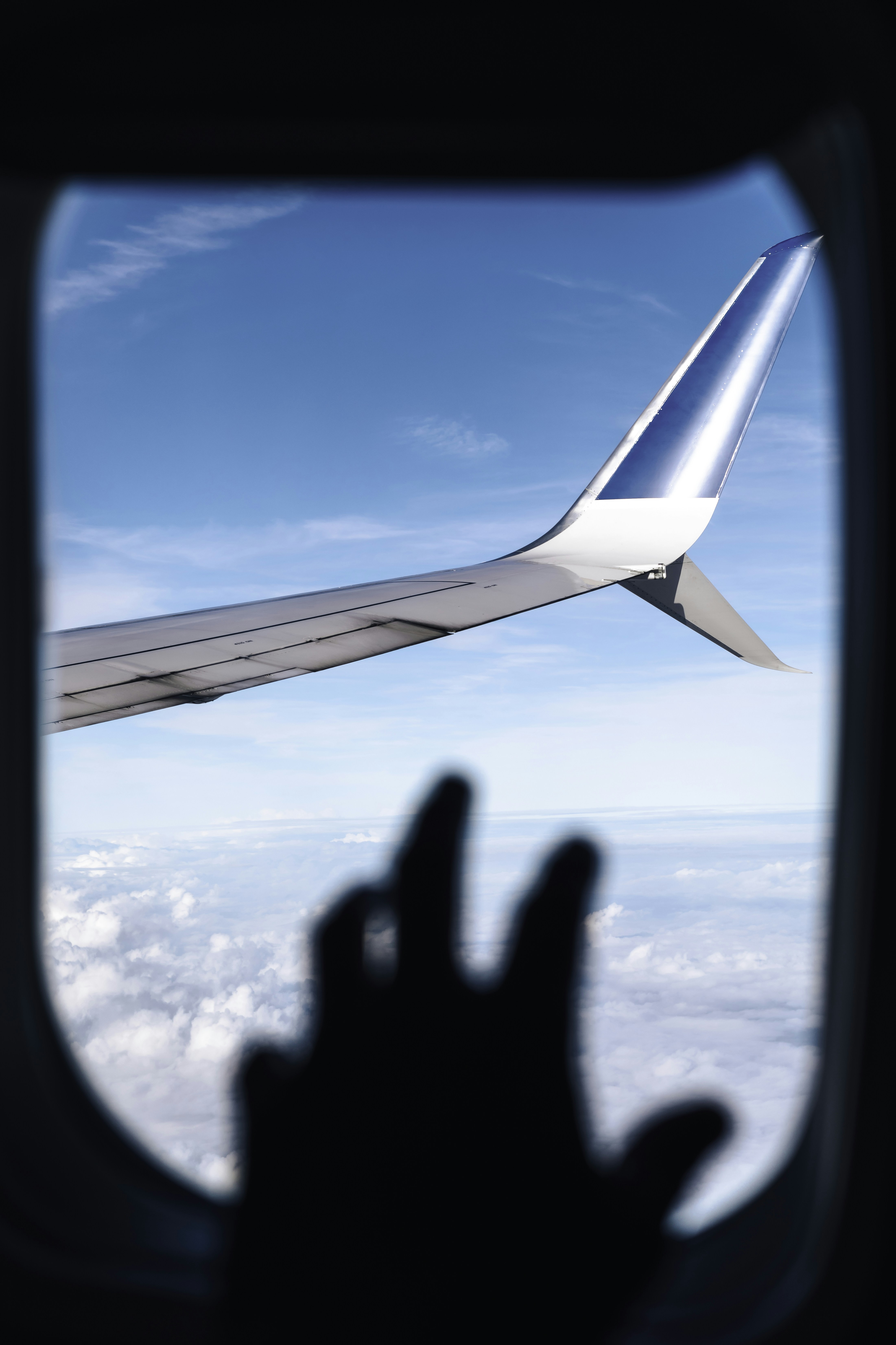 Silhouette of a hand reaching towards the airplane wing through a window, framed by a serene sky filled with clouds.