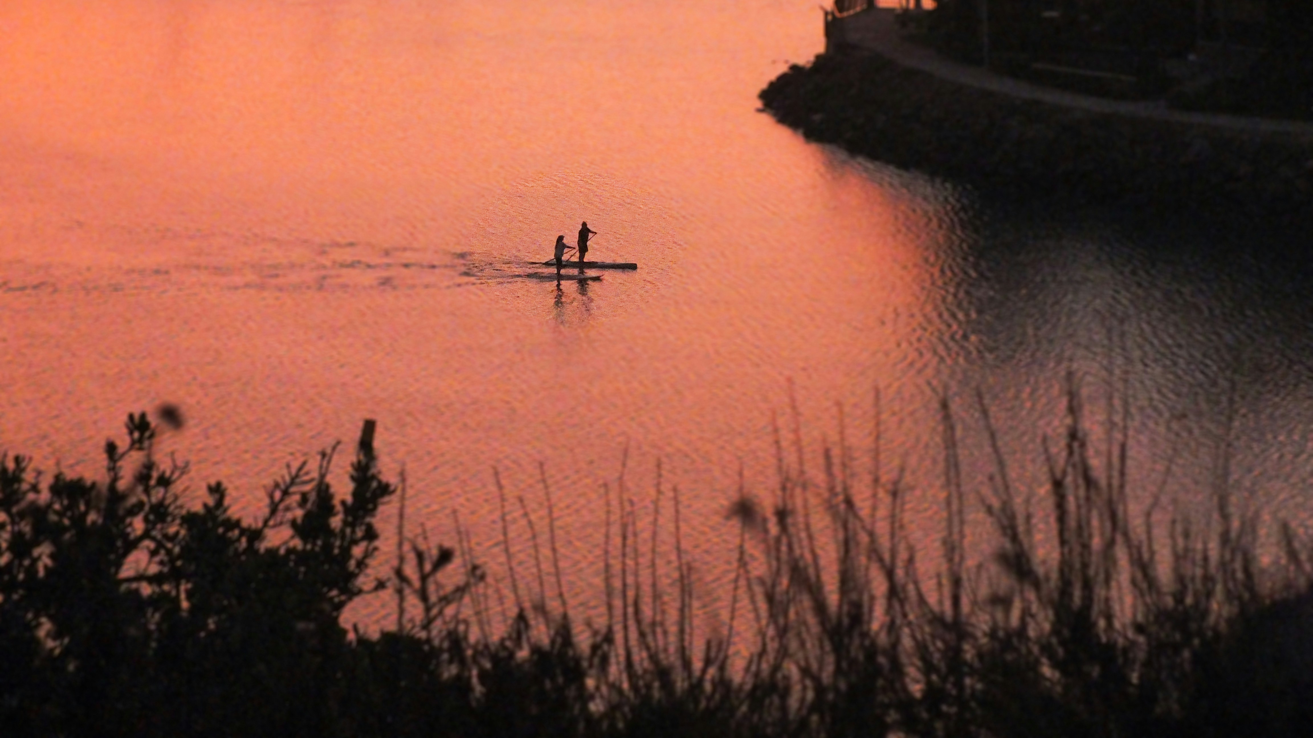 Two figures paddle across a tranquil body of water at dusk, framed by soft hues of pink and orange. The scene captures the peaceful essence of twilight.