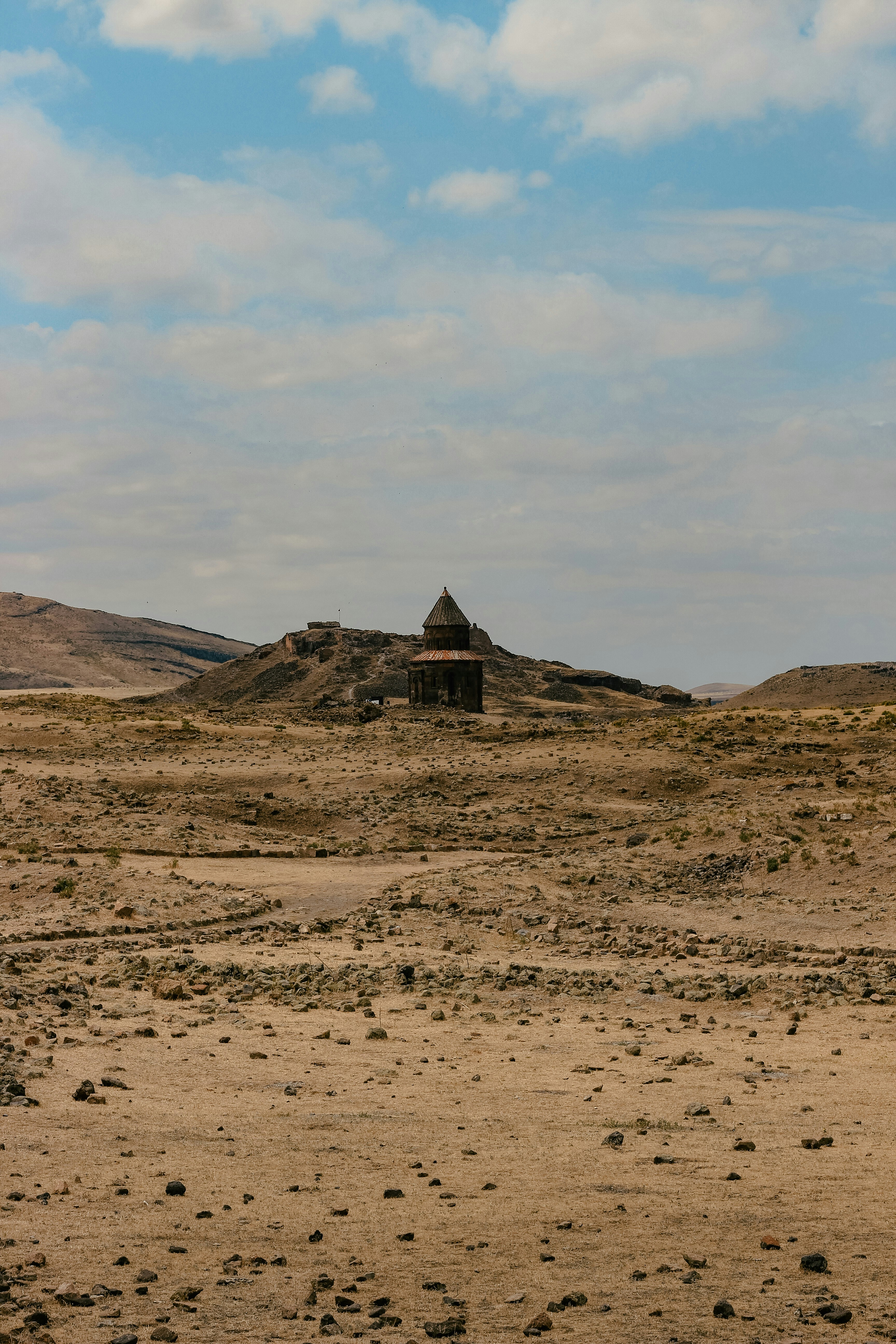 Ancient structure stands alone in a vast, arid landscape, surrounded by scattered stones and distant hills. A testament to time and resilience.