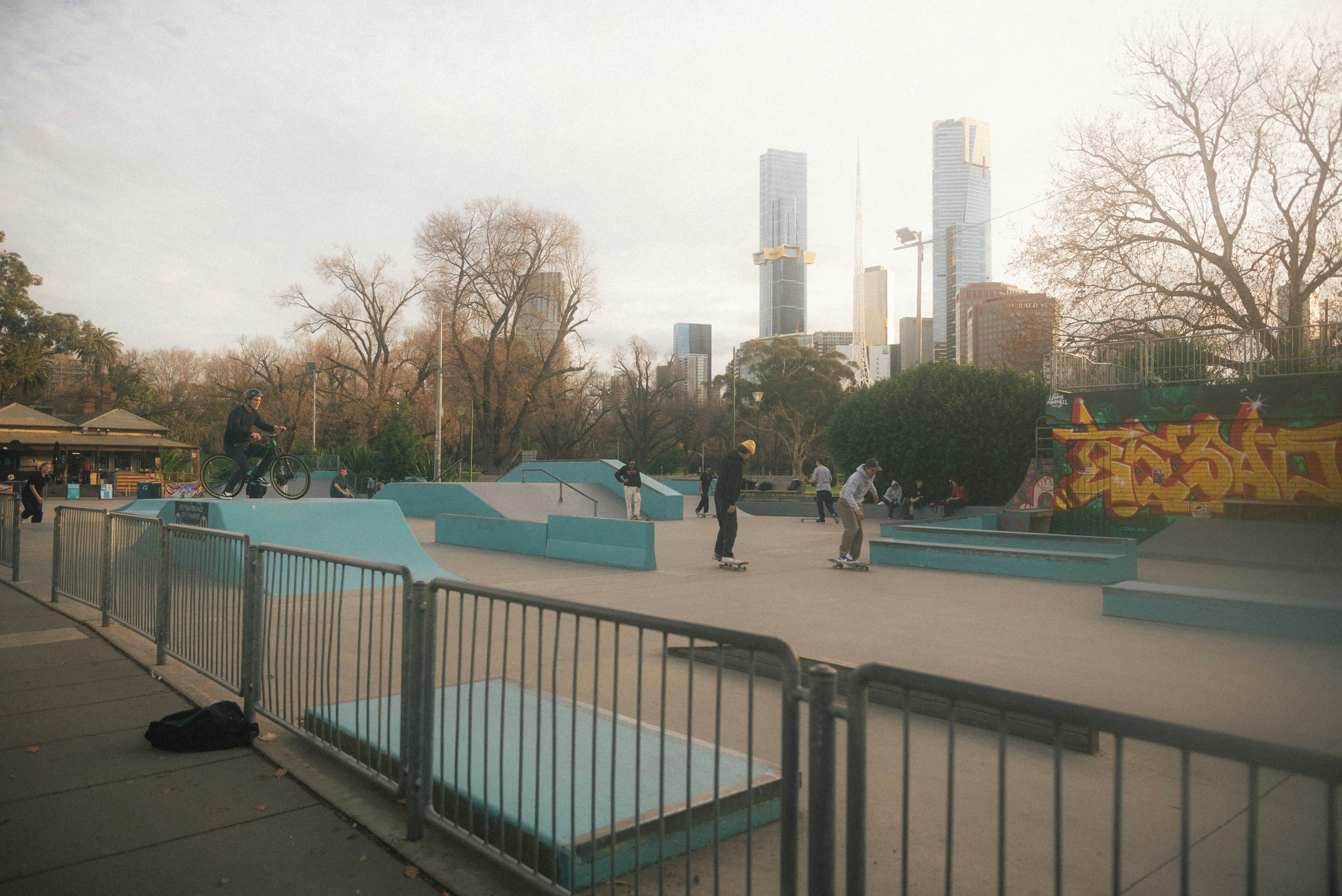 Skateboarders showcasing tricks in a vibrant urban skate park, framed by city skyscrapers and autumn trees.