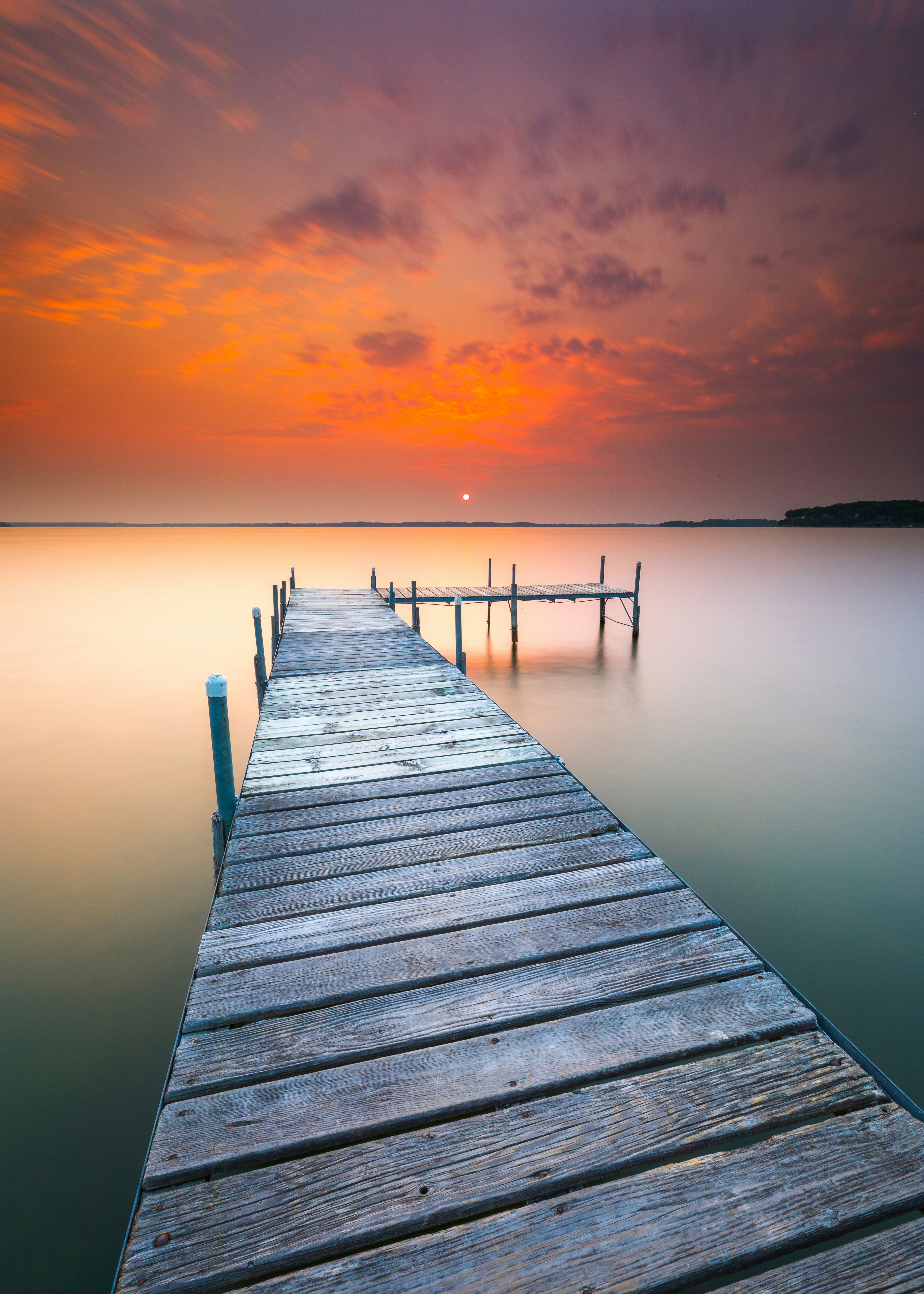 Wooden pier extending into a serene lake under a vibrant sunset sky with wispy clouds.