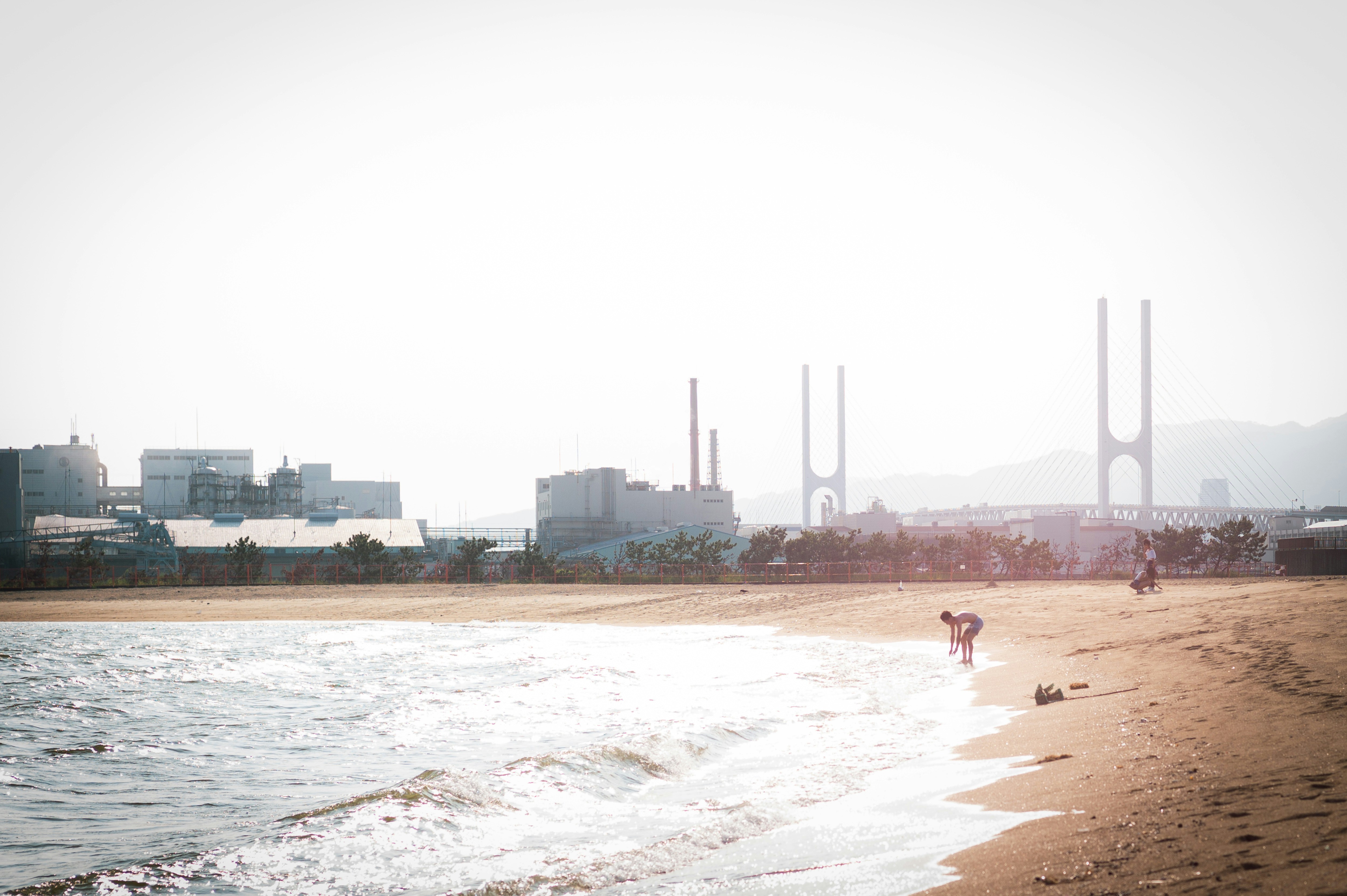 Sandy beach with gentle waves under an industrial skyline and distant mountains.