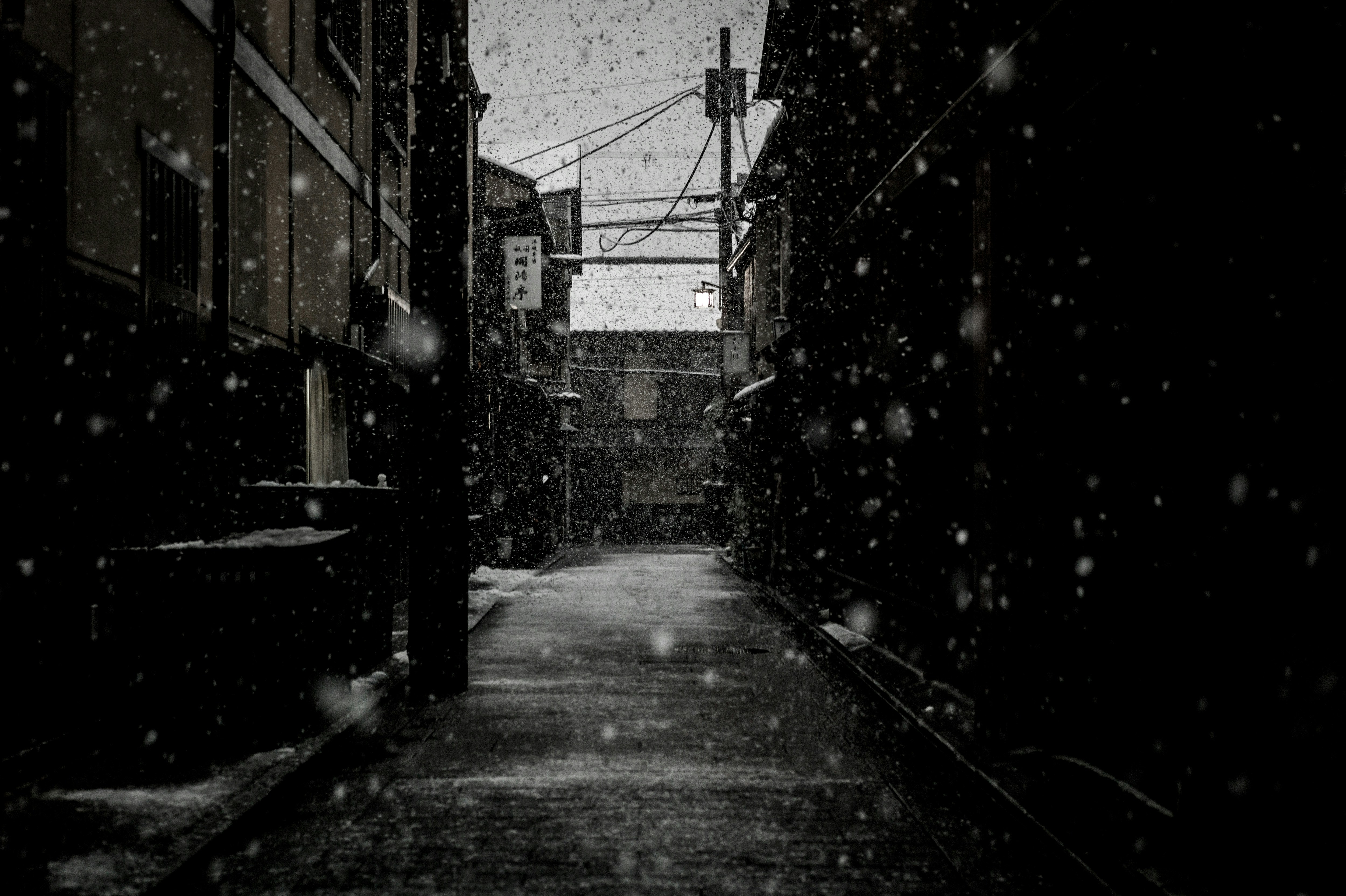 Black-and-white photograph of a narrow alley receding toward a faint light, with snow flurries accenting the mood.