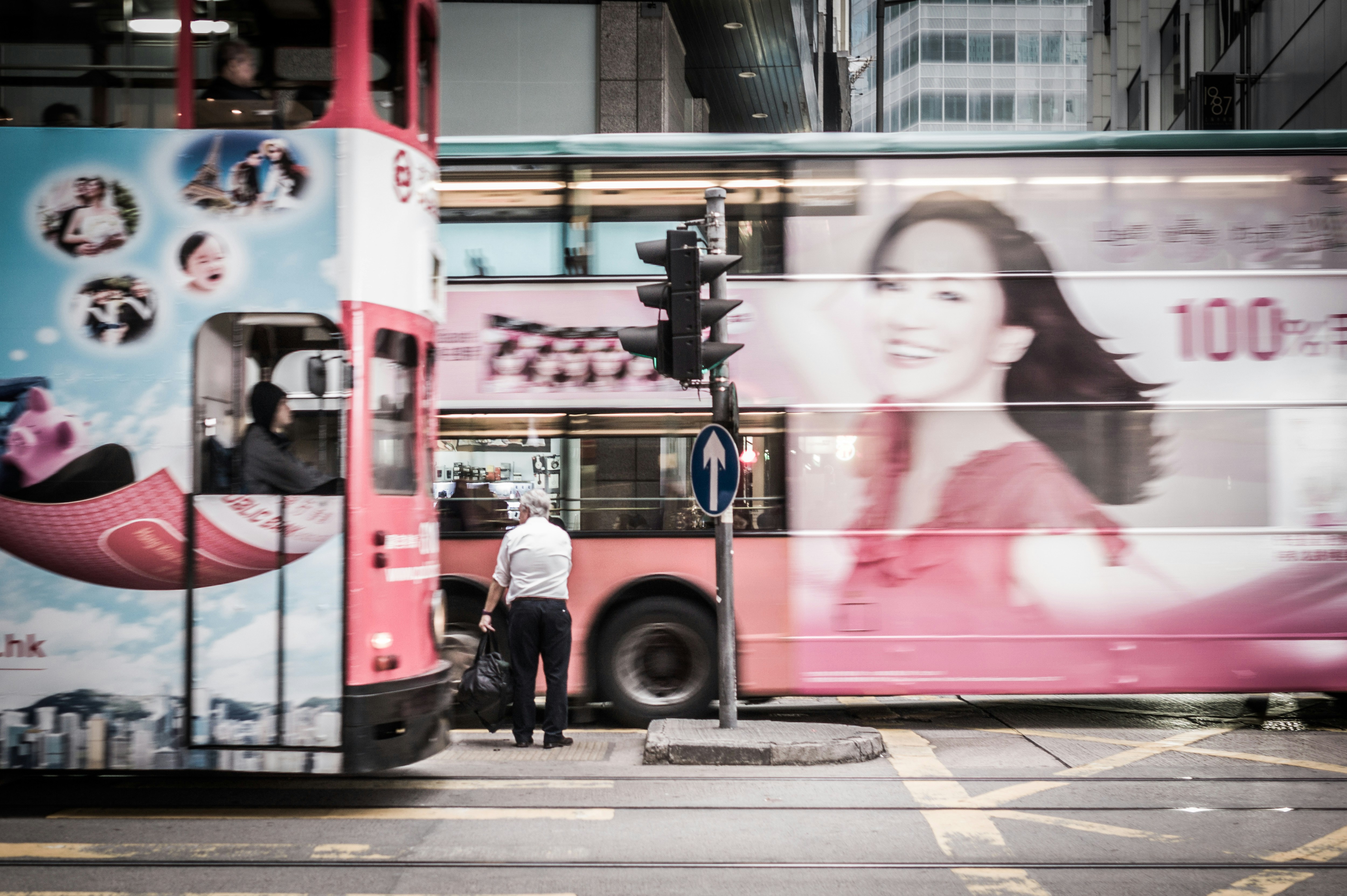 Urban street photograph showing a blurred advertising bus passing behind a lone pedestrian at a crosswalk, framed by a traffic signal.