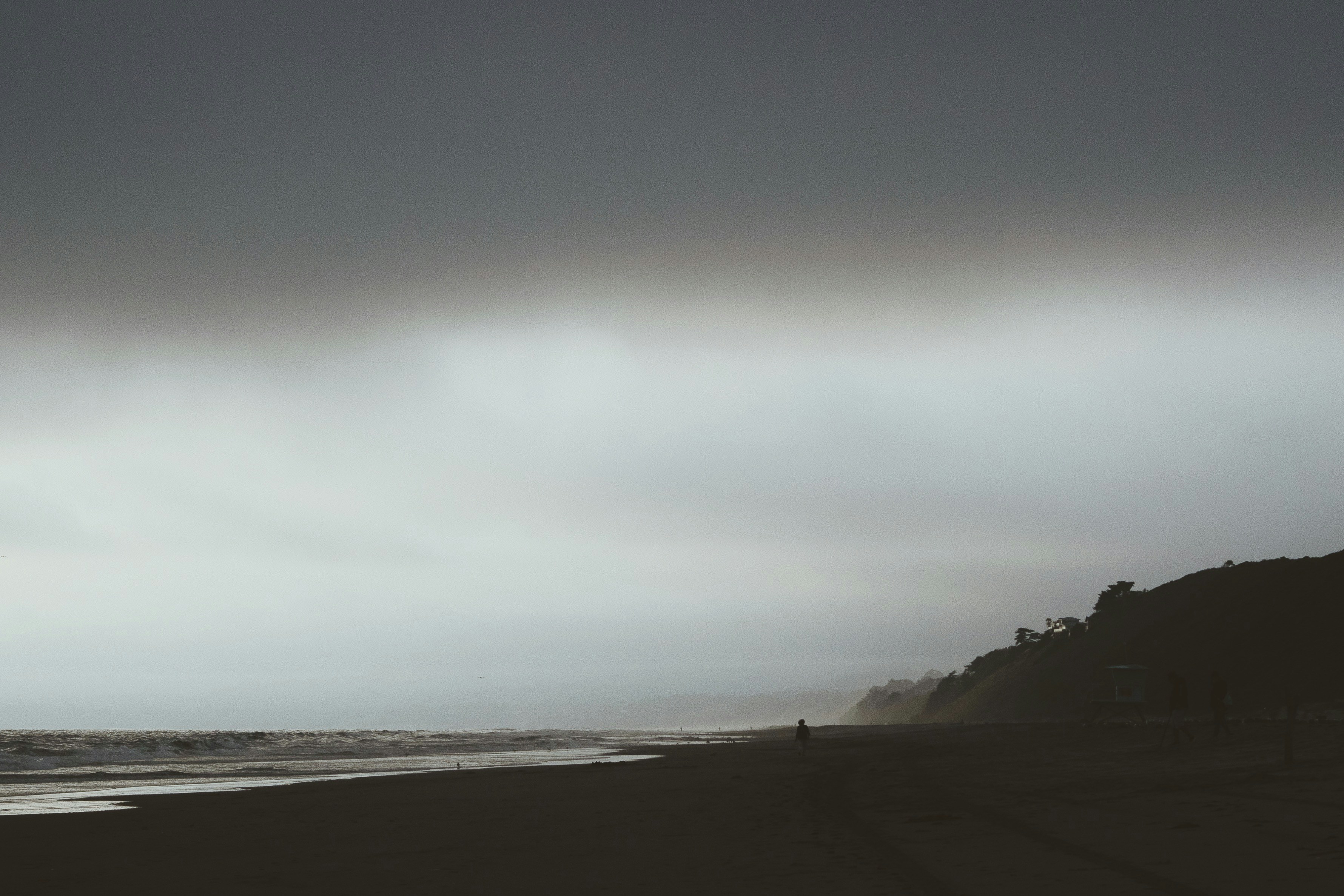 A lone figure strolls along a desolate beach under a dramatic, overcast sky. The muted tones create a serene atmosphere.