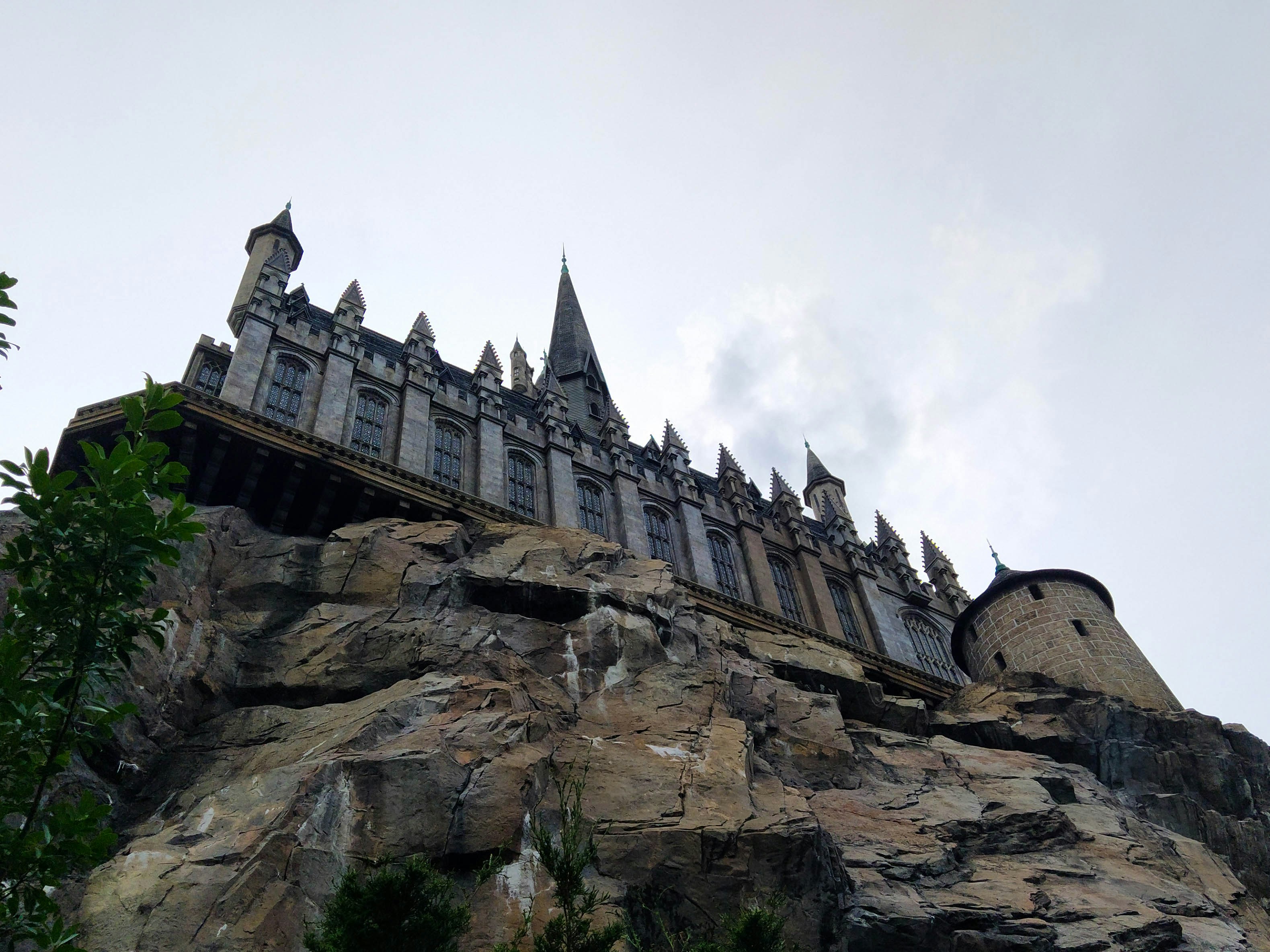 Gothic castle perched atop a rocky outcrop, surrounded by lush greenery and a cloudy sky.