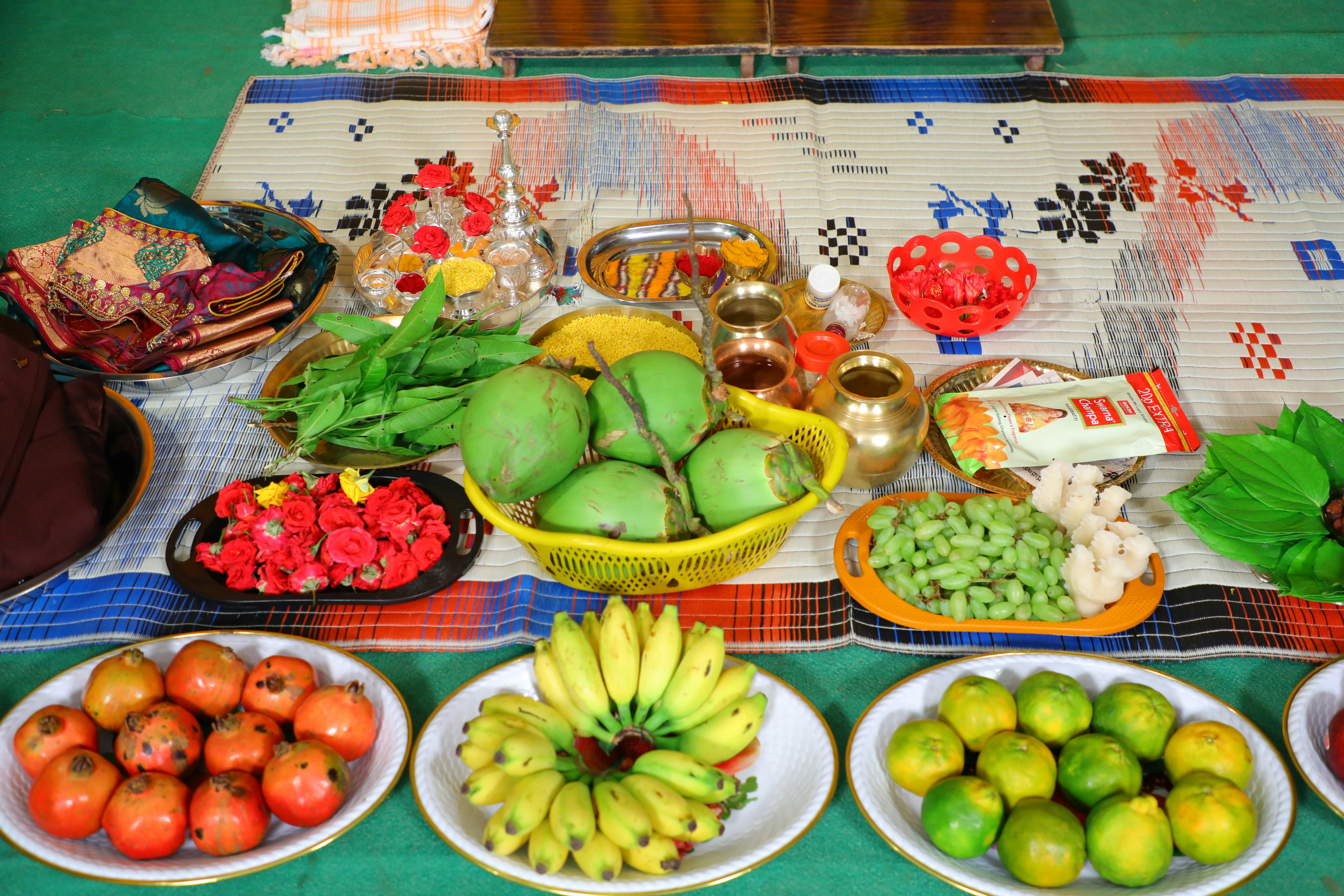 A colorful assortment of fruits, herbs, and decorative items arranged on a traditional mat, showcasing a cultural celebration. The scene reflects a rich tapestry of textures and colors.