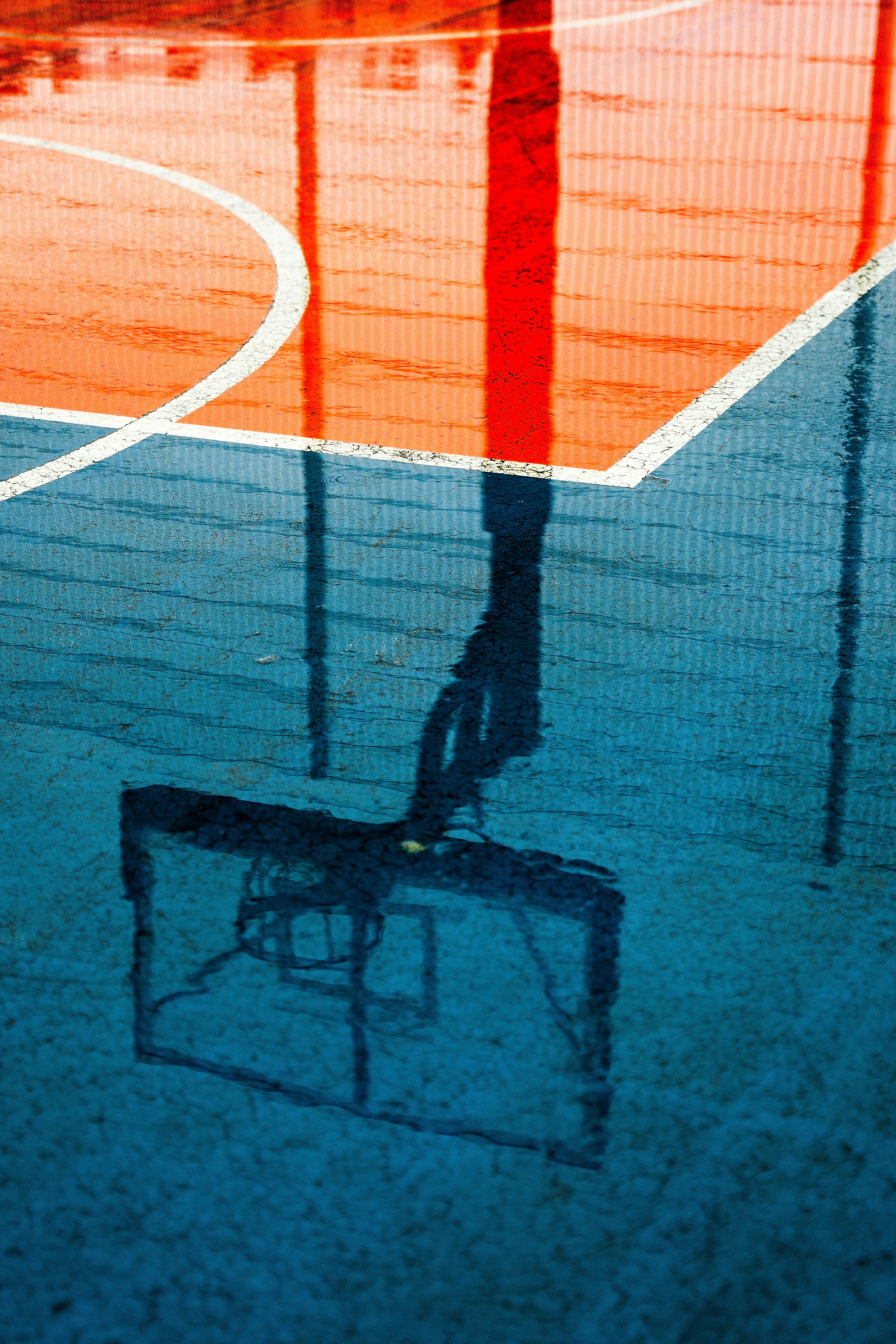 Vibrant basketball court reflecting the hoop and surrounding lines in a pool of water. The contrast of orange and blue creates a striking visual.